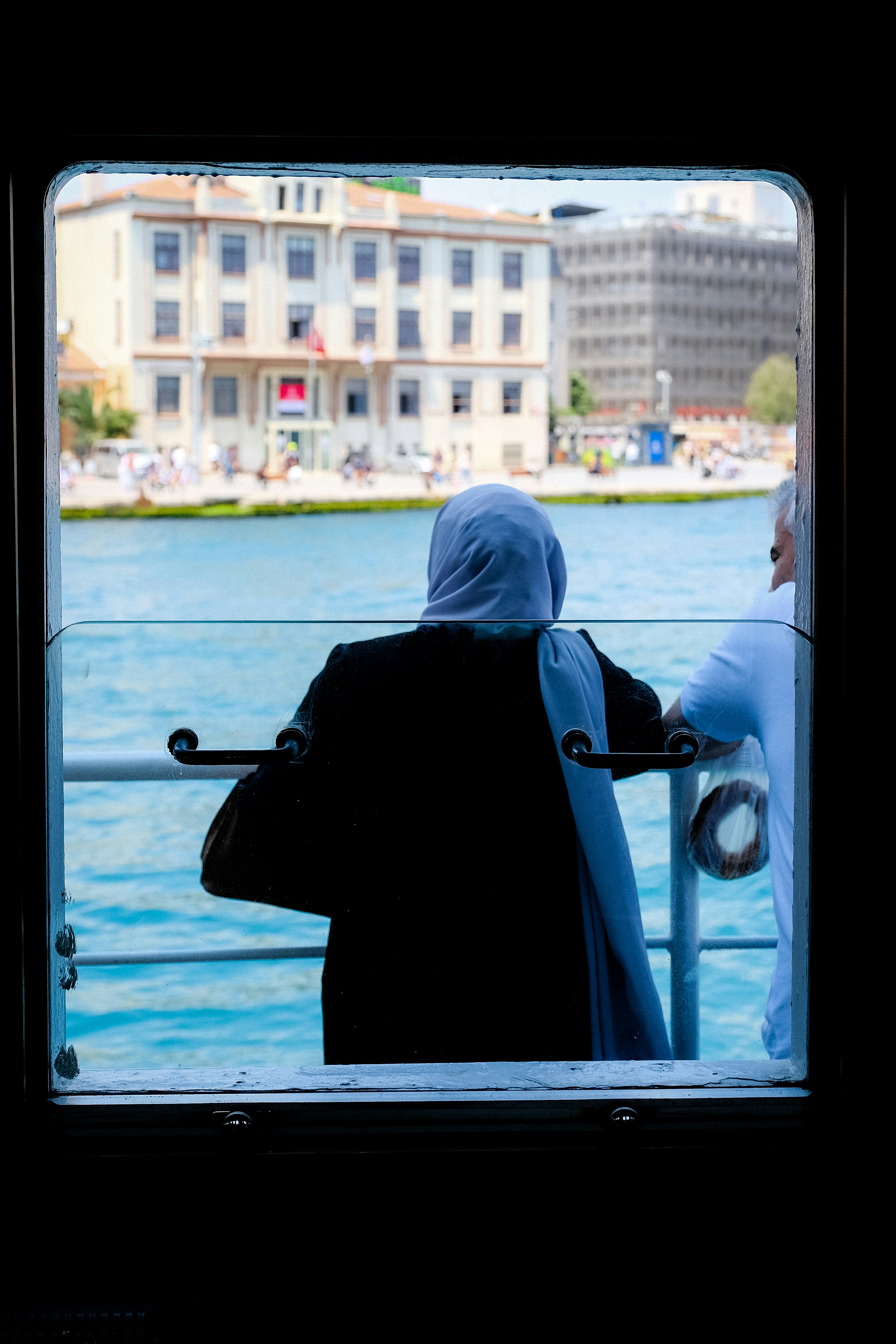 A woman in a black hijab gazes out at the waterfront, framed by a boat window, while another figure stands beside her. The scene captures a moment of contemplation amidst a vibrant backdrop.