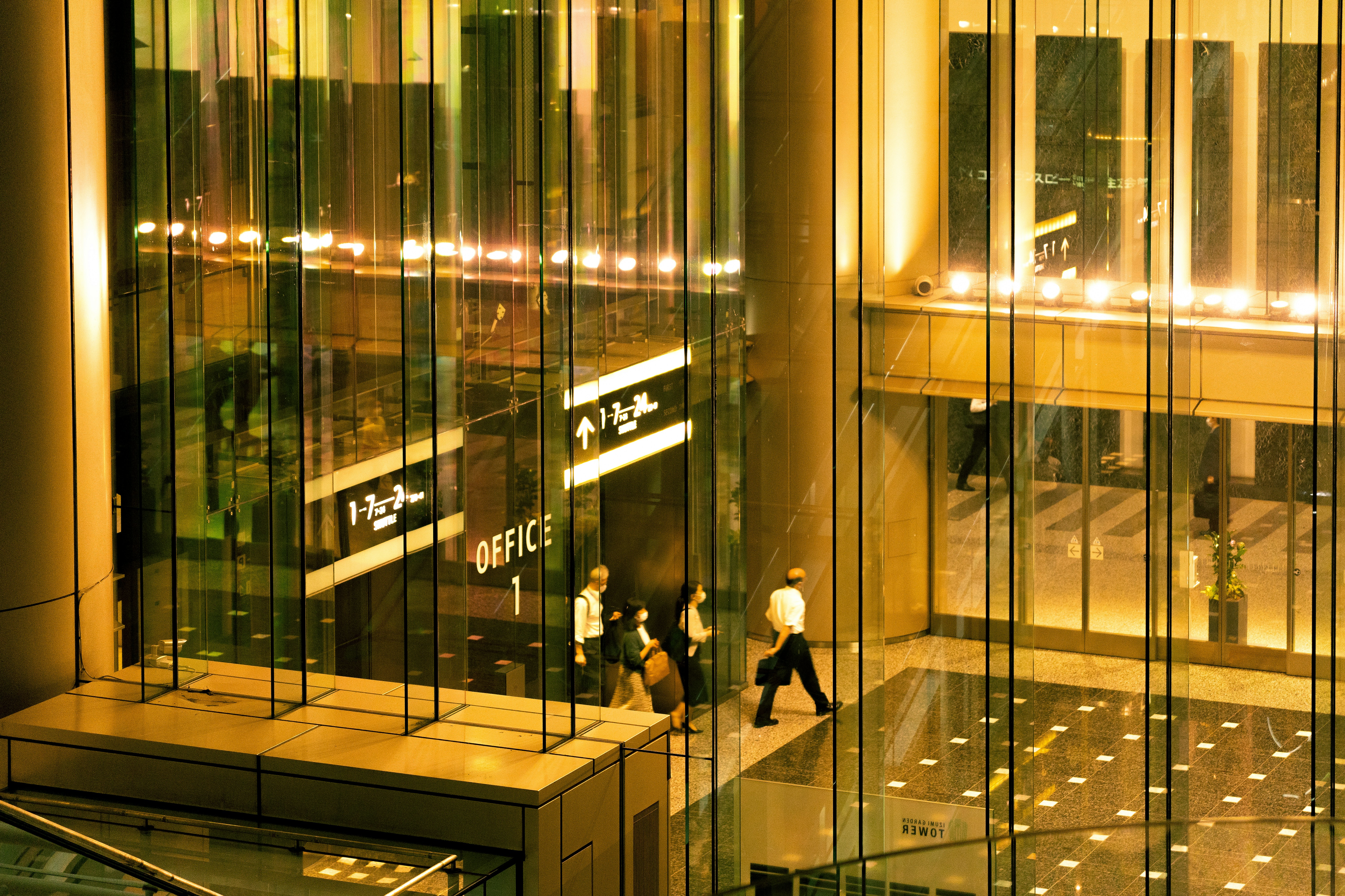 Business professionals walking past illuminated office signage in a modern glass building. The reflective surfaces create a dynamic interplay of light and shadow.