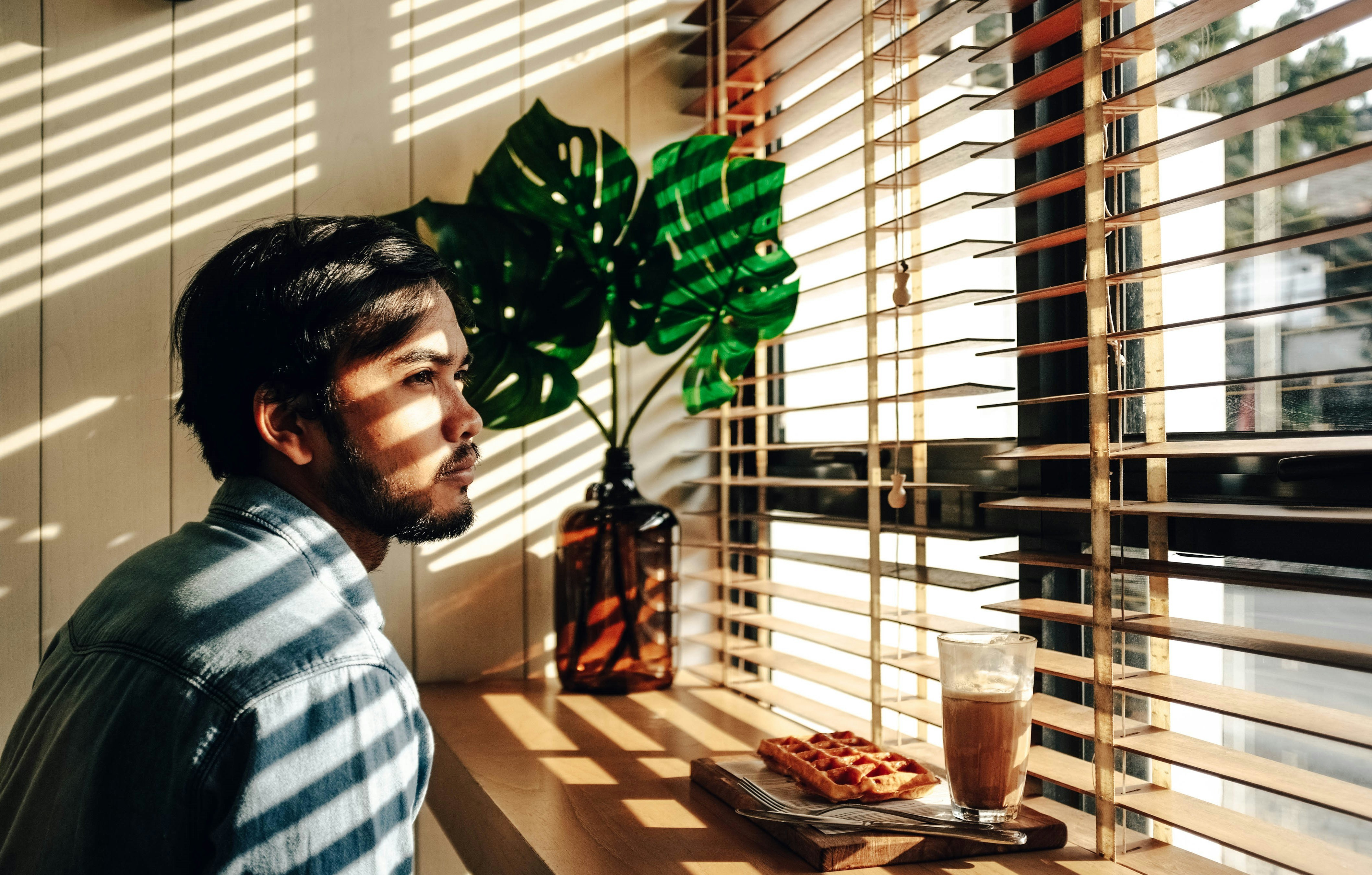 A contemplative man gazes out the window, bathed in patterned sunlight, with a plate of waffles and a glass of iced coffee nearby.