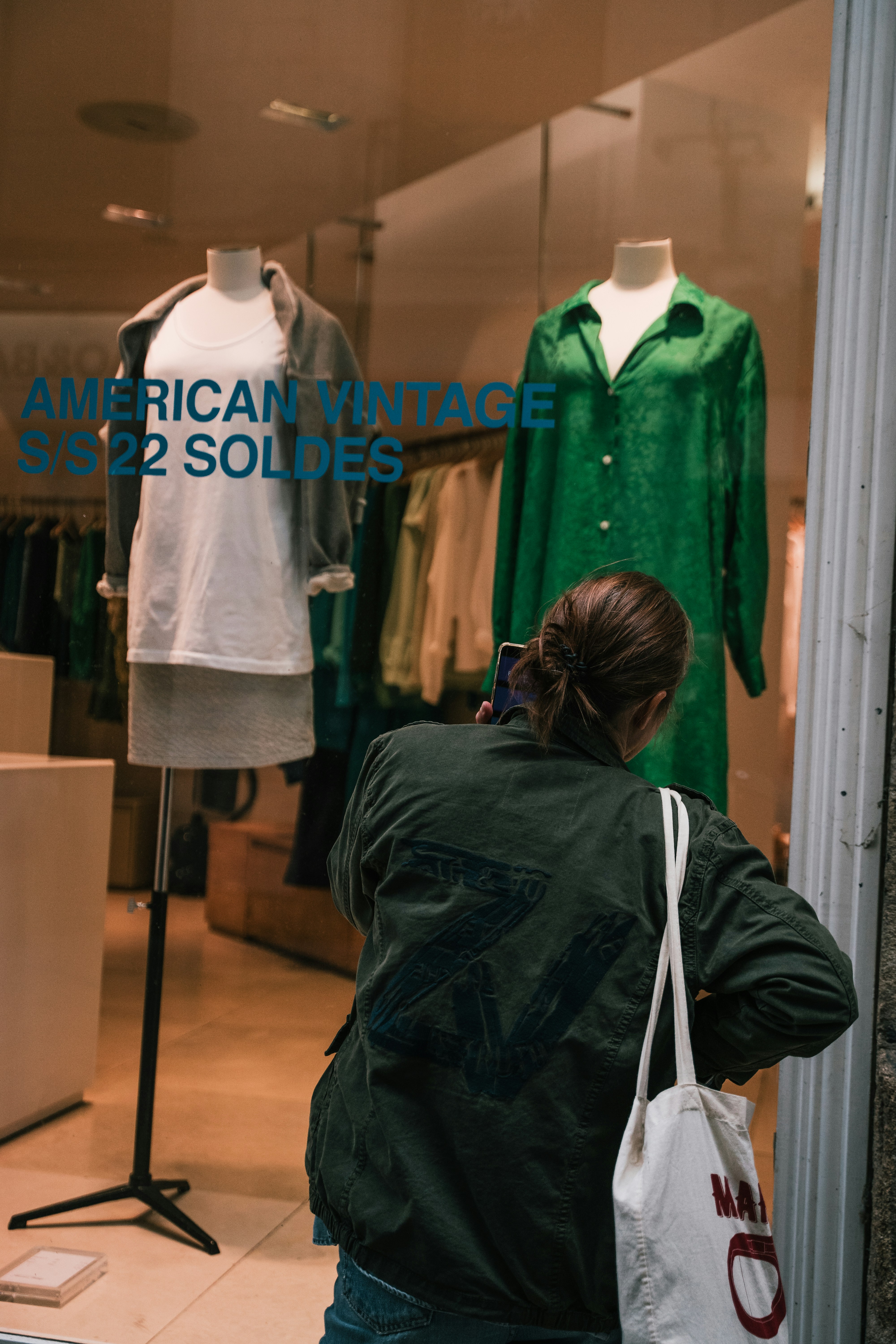 A shopper admires vintage clothing displayed in a boutique window, featuring a green shirt and a casual white outfit. The store's promotional text is prominently featured.