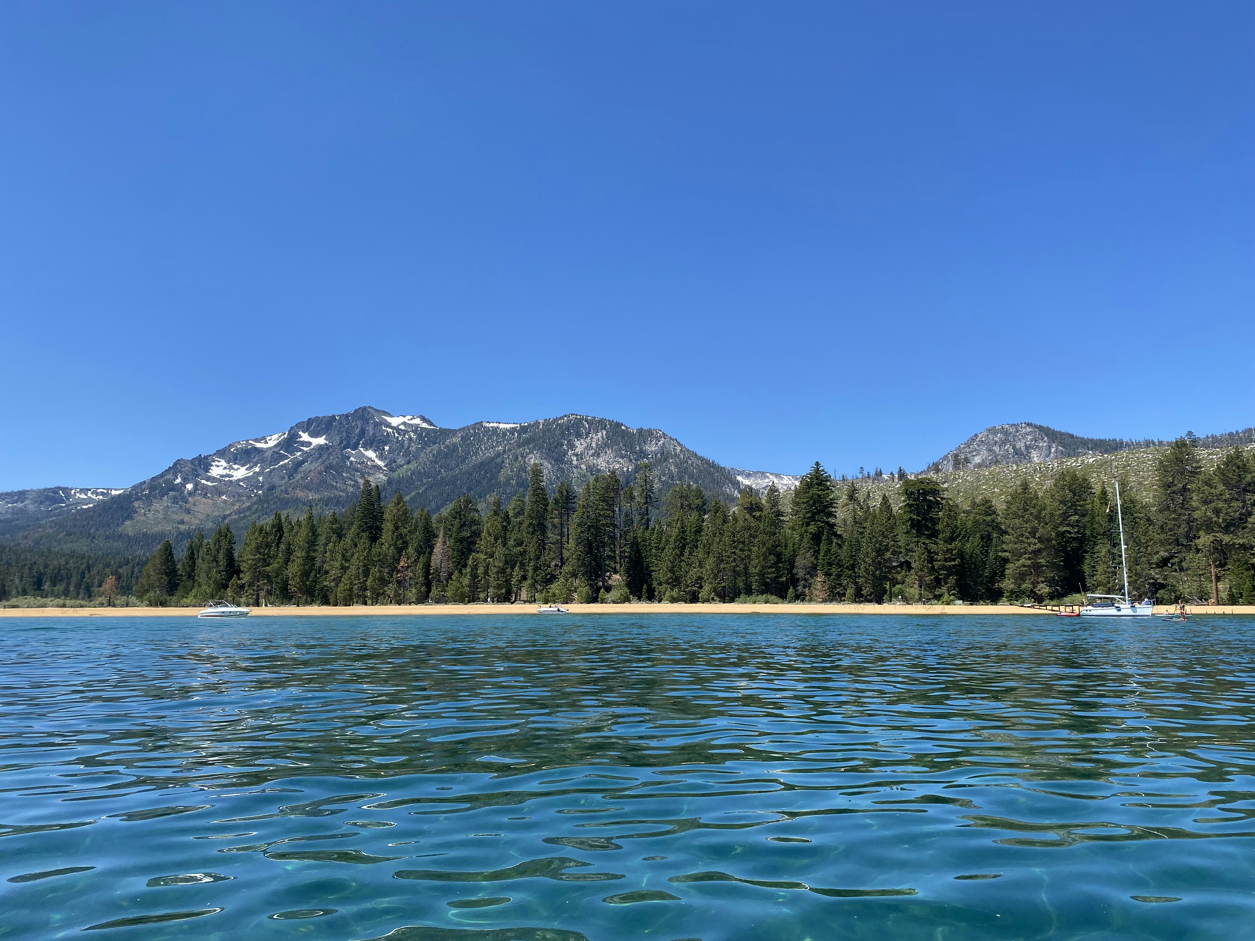 Clear blue lake reflecting distant mountains and a line of trees under a bright sky.