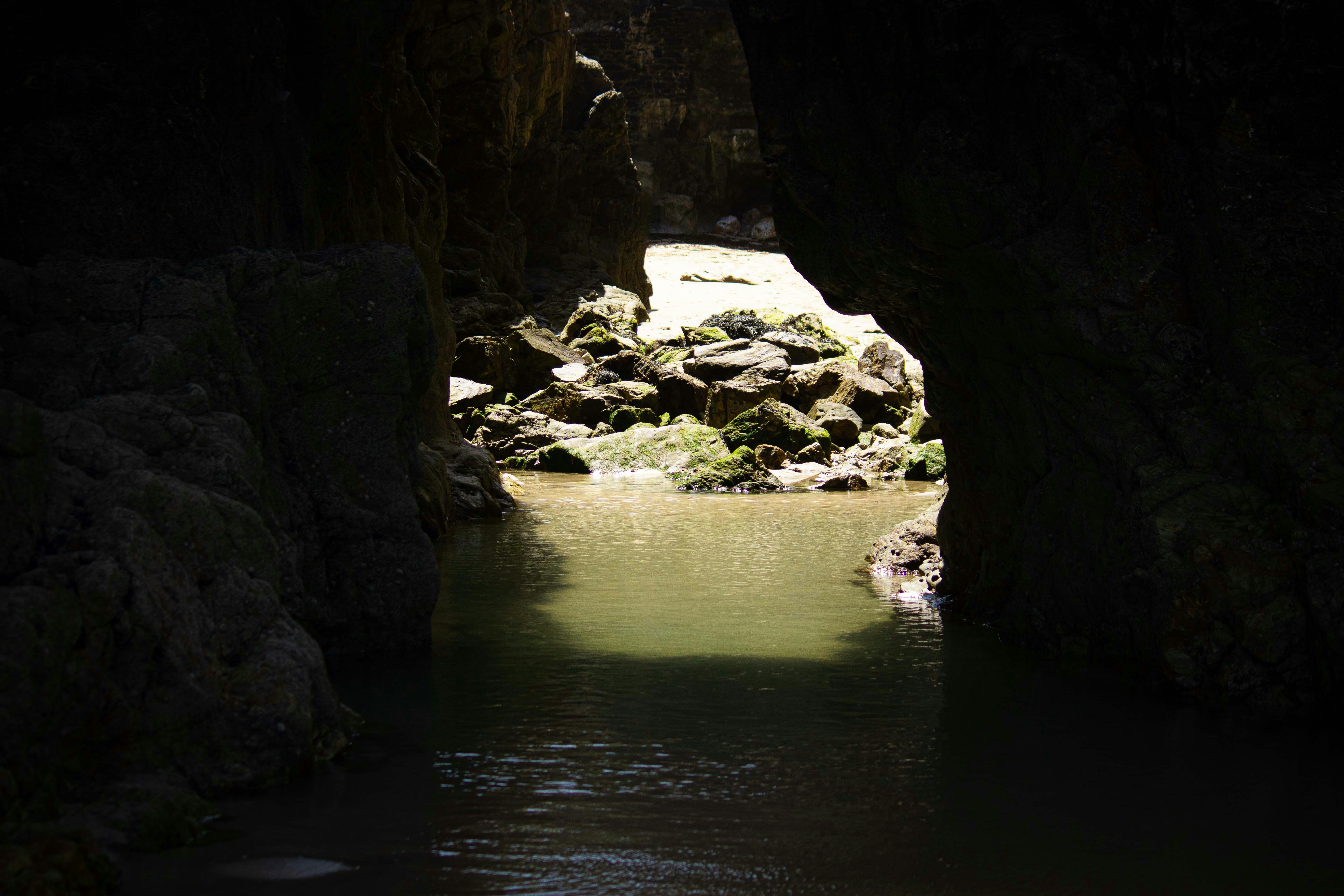 Sunlight filters through a rocky archway, illuminating a serene pool surrounded by moss-covered stones. The contrast between light and shadow creates a tranquil atmosphere.