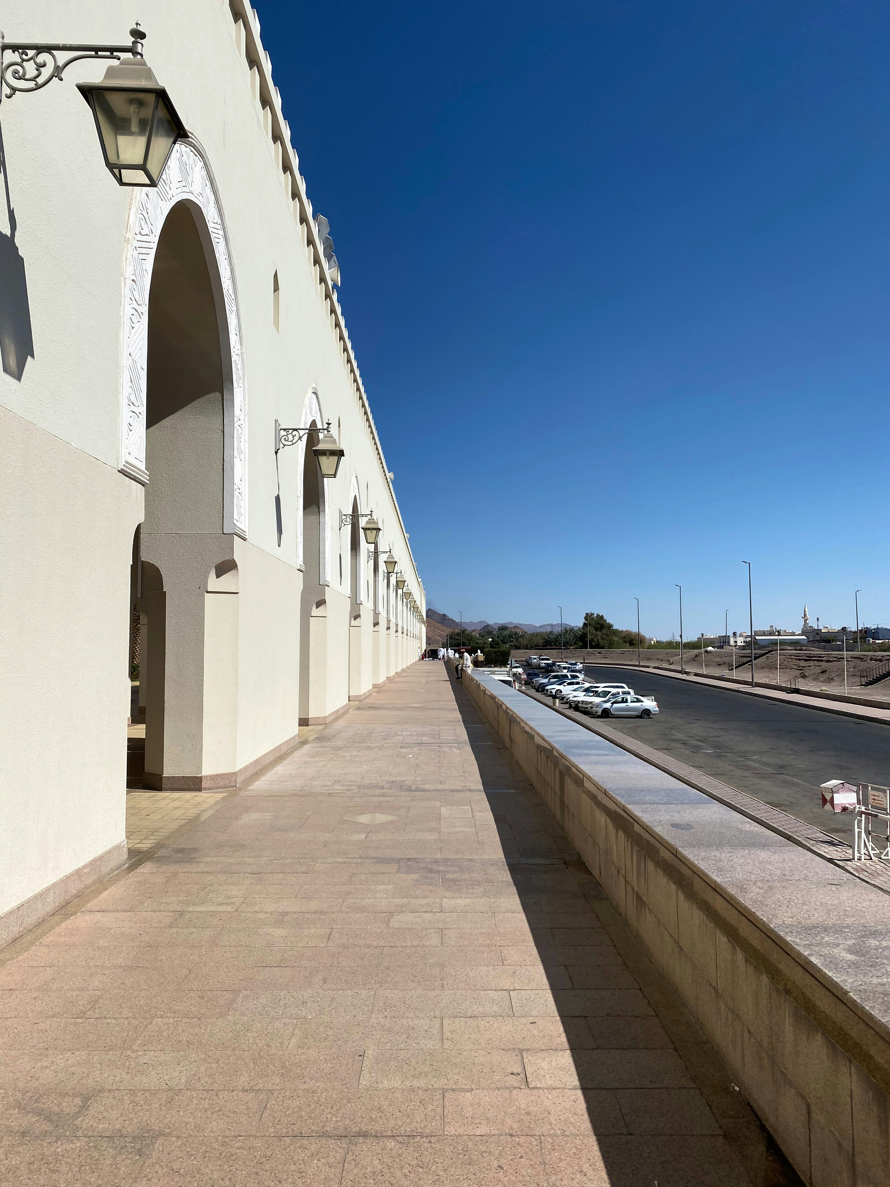 Long walkway lined with arches and lamps, leading to a distant road with parked cars under a clear blue sky.