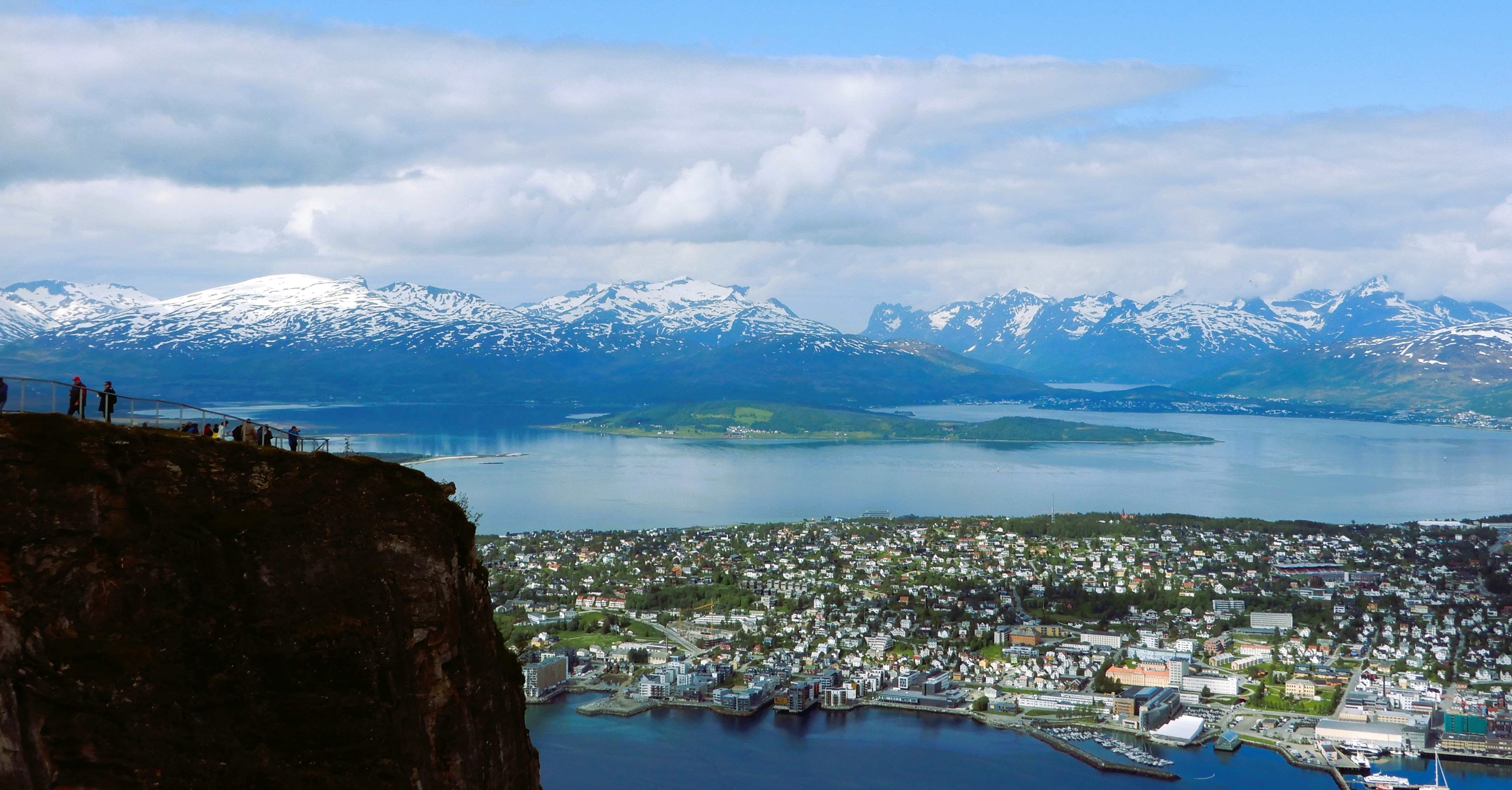 Dramatic cliff overlooking a vibrant coastal town with snow-capped mountains in the background, showcasing the beauty of nature's contrasts.