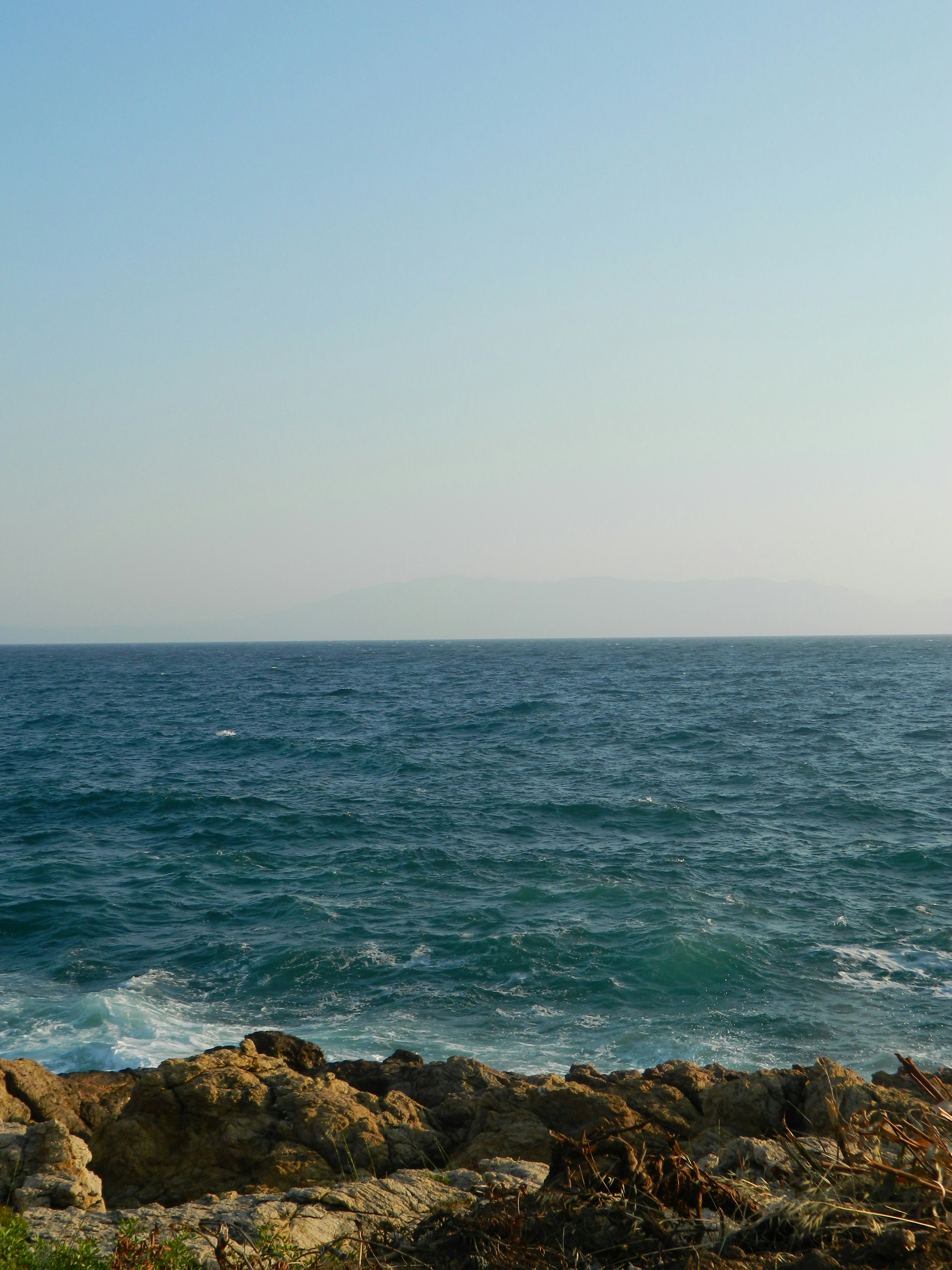 Waves crash against rocky shoreline under a clear sky, with distant hills fading into the horizon. 