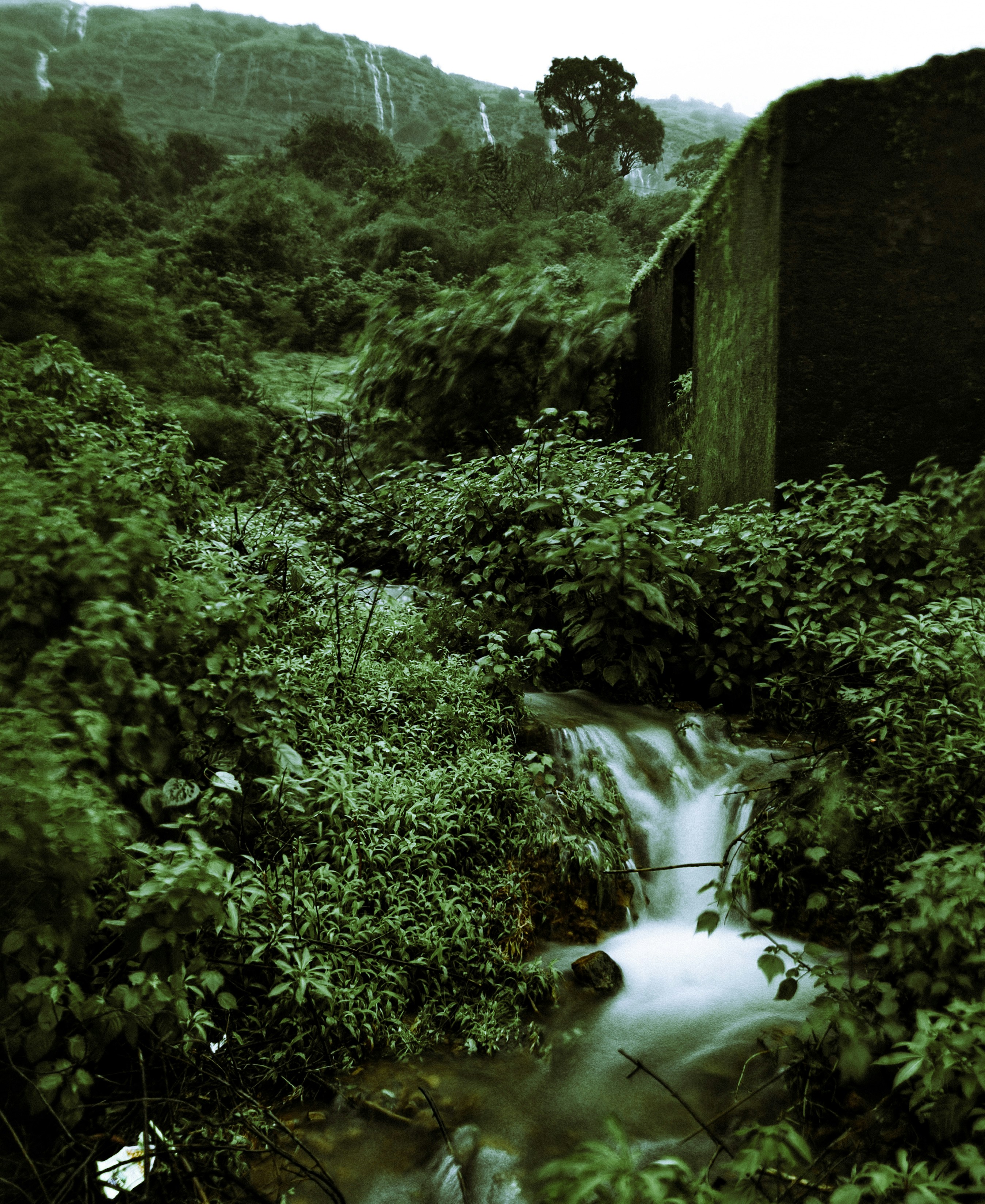 A serene stream flows through lush greenery beside a weathered structure, with distant waterfalls cascading down the hills. 