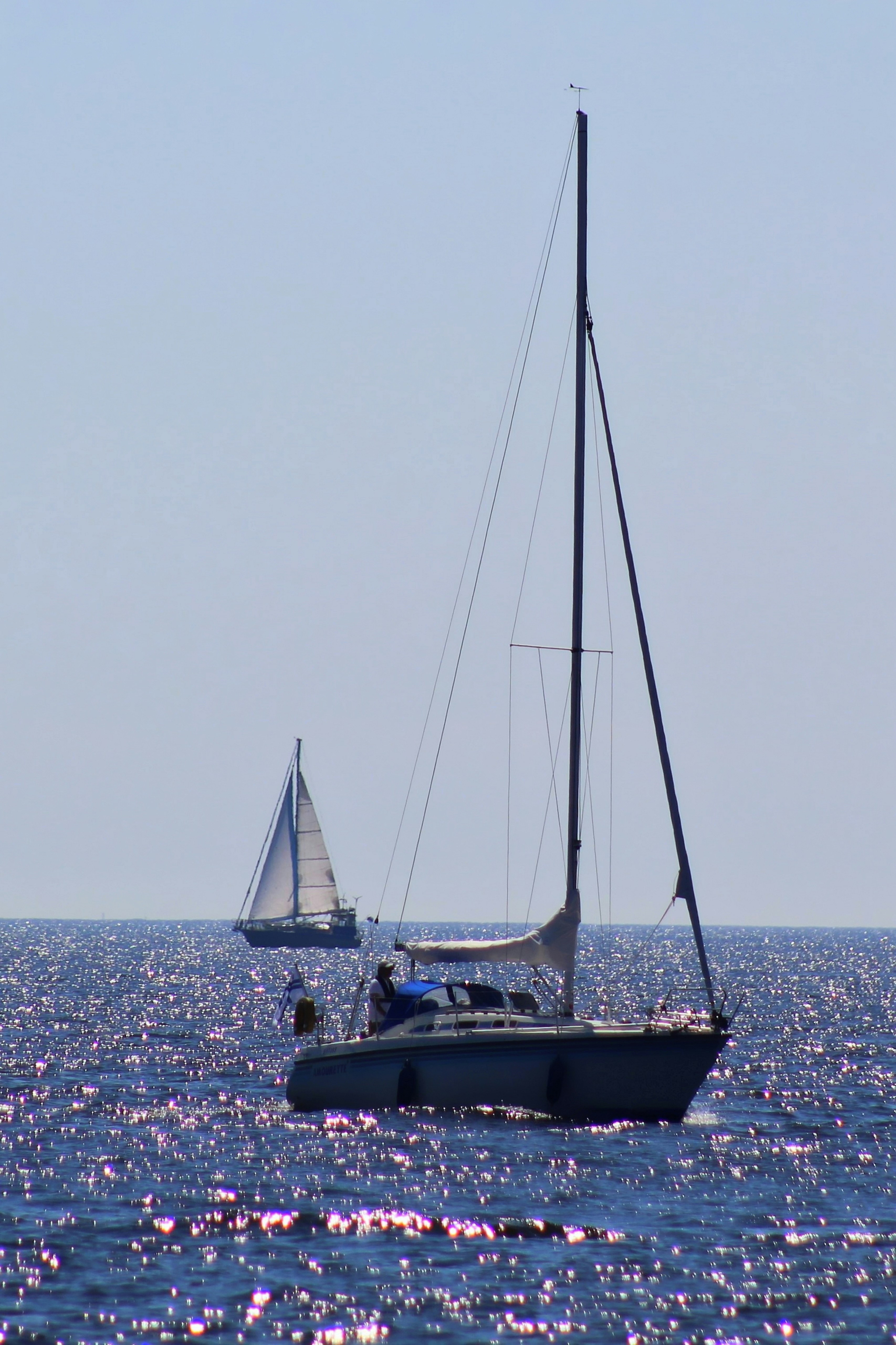 A sailboat gliding across shimmering ocean waves, with another sailboat in the background under a clear sky.