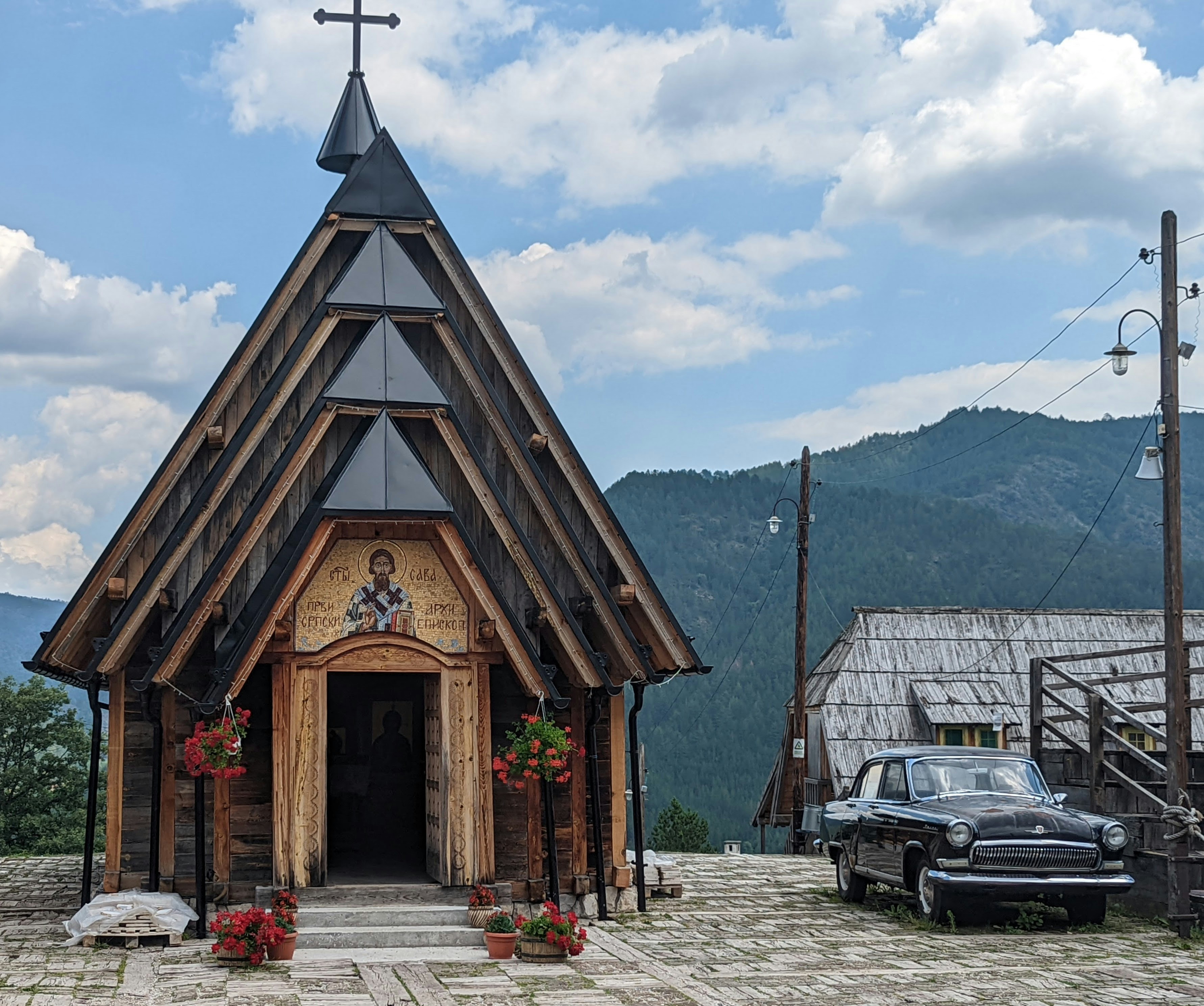 Wooden chapel with a triangular roof next to a vintage car, set against a mountainous backdrop and cloudy sky.