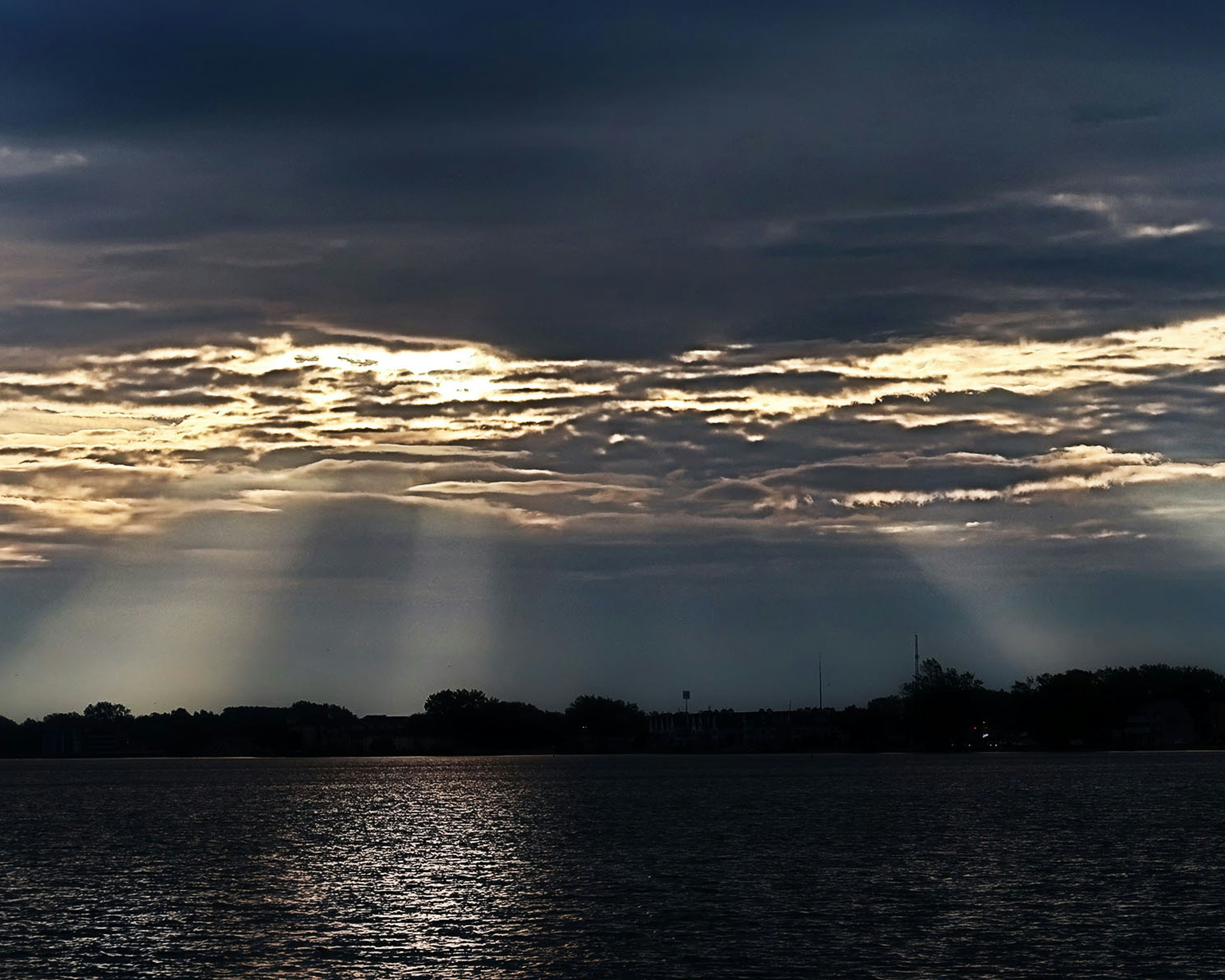 Sunbeams breaking through cloudy skies over a calm body of water, with silhouetted treetops on the horizon.