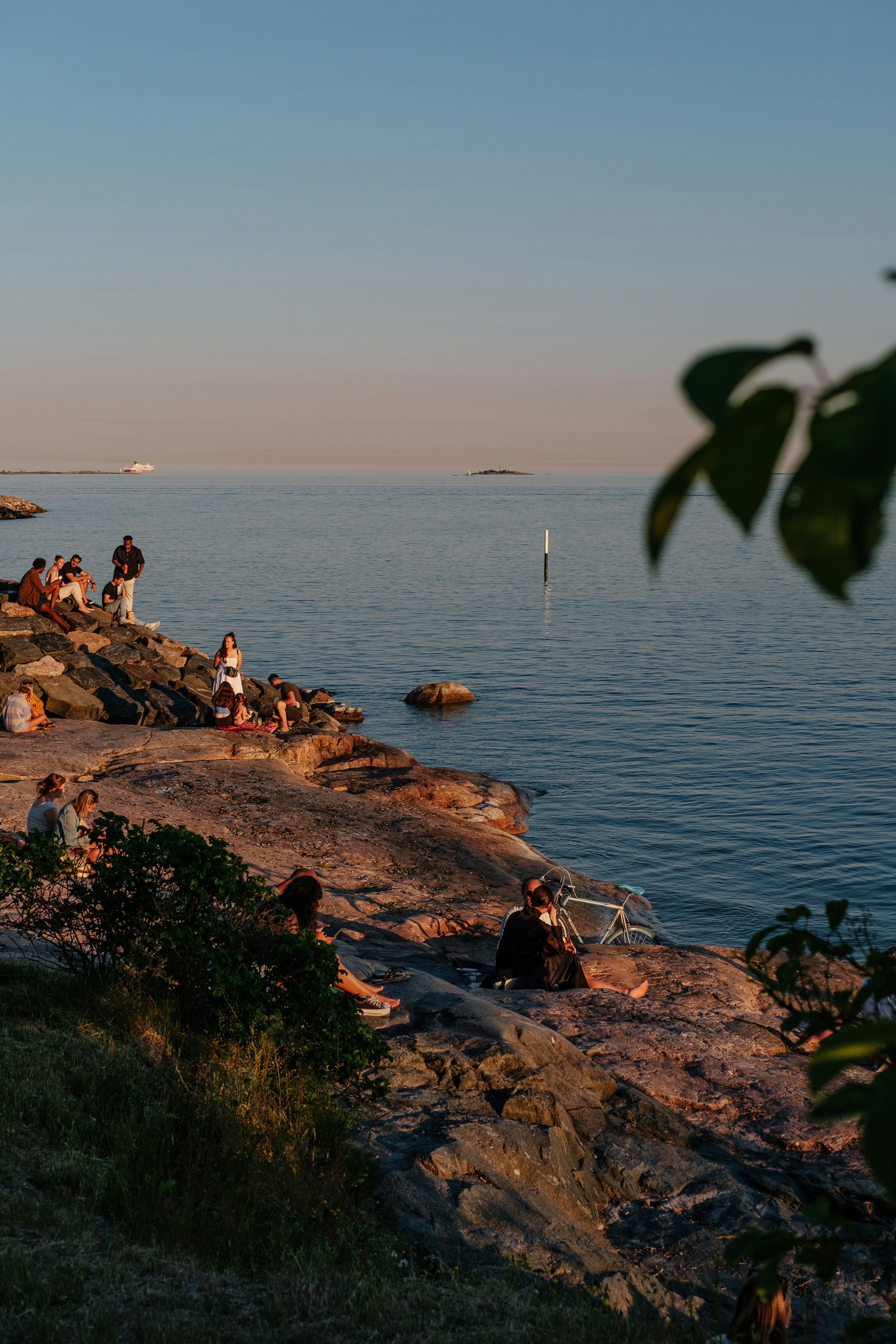 People gather on rocky shoreline at sunset, enjoying the tranquil waters and fading light. The scene captures a peaceful moment by the sea.