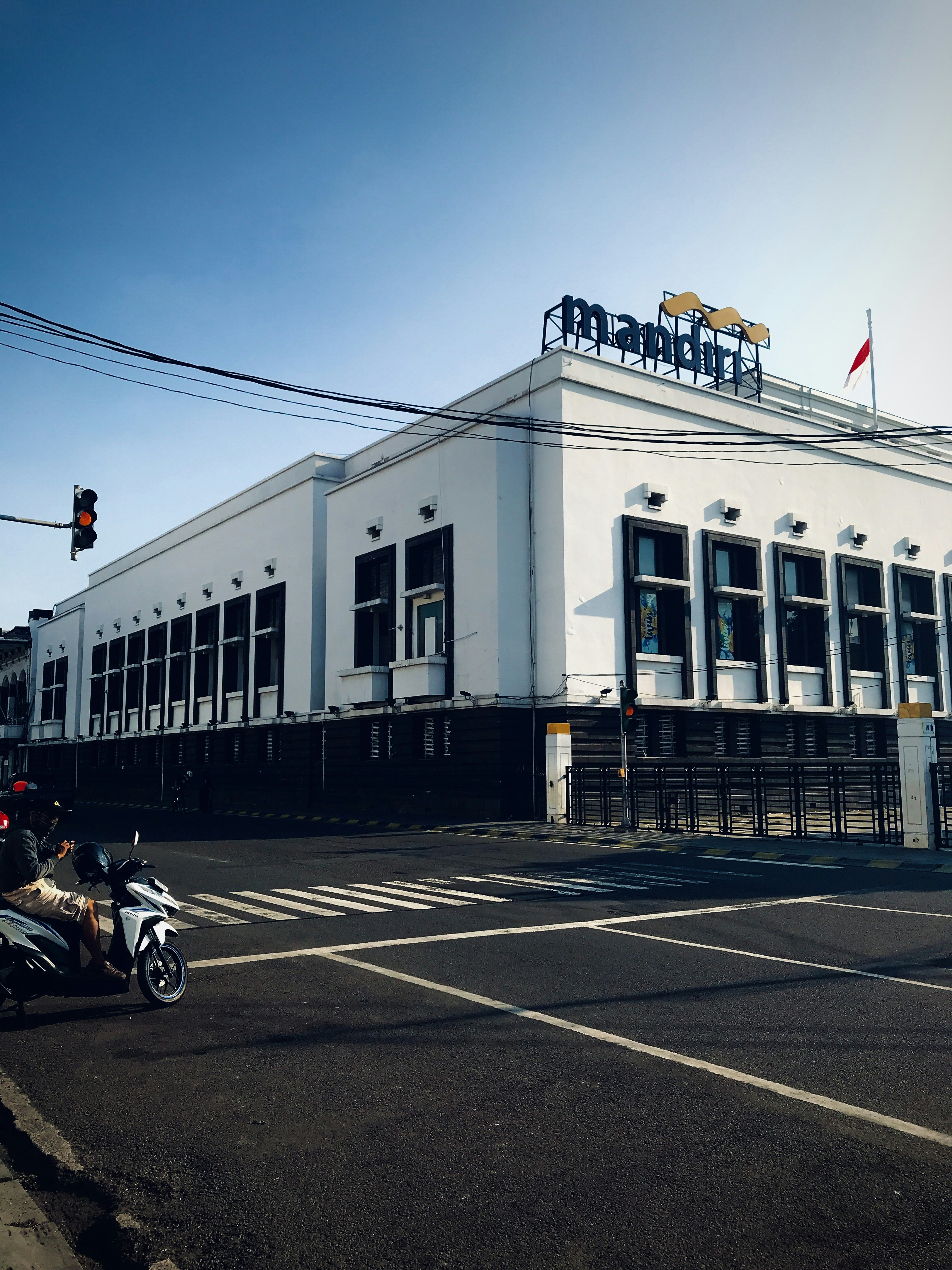 A quiet street scene where a pale blue building with a MANDIRI sign looms over a crosswalk and parked scooter at a sunlit intersection