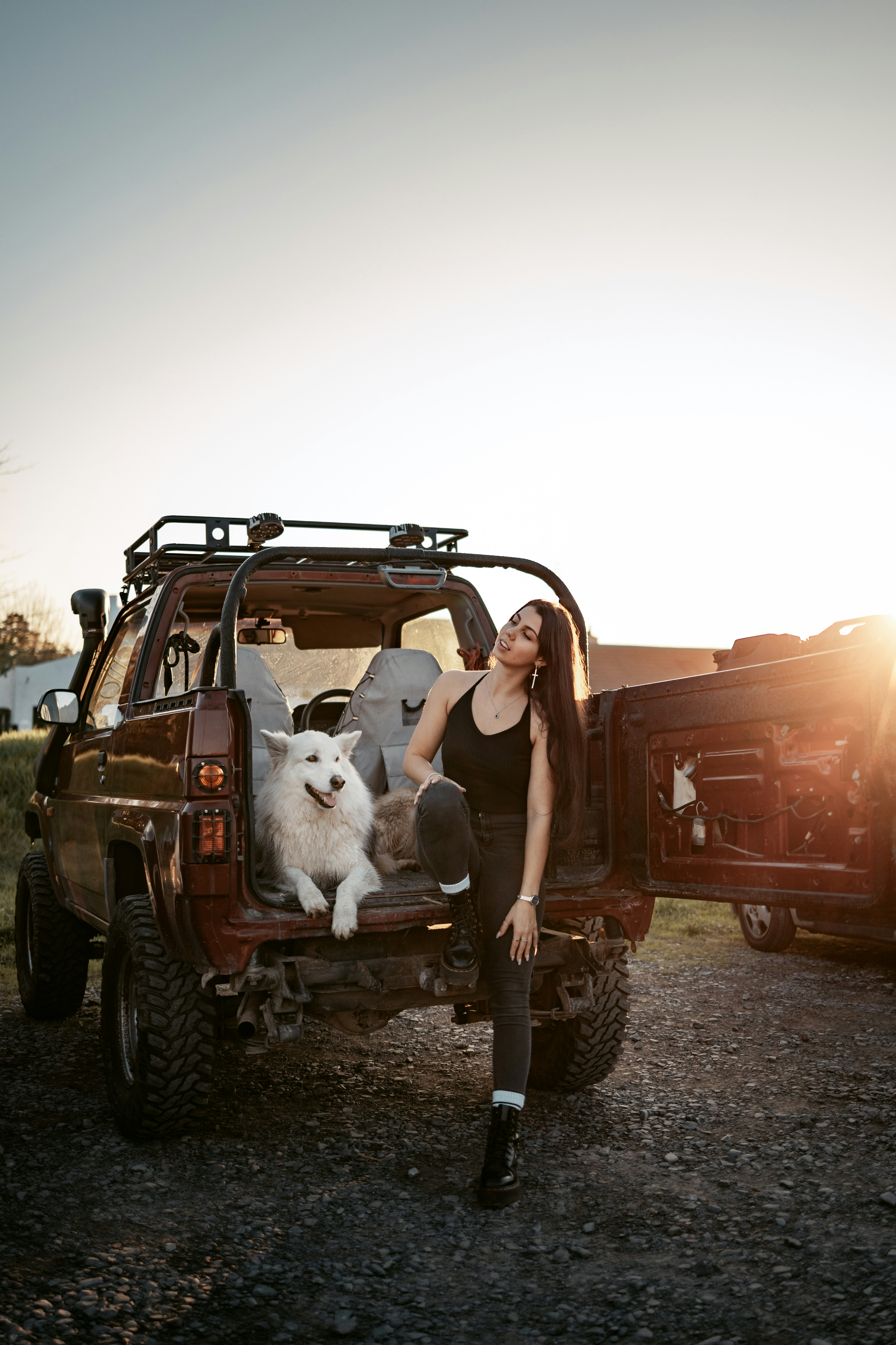 Woman lounging in the back of an SUV with a fluffy white dog, basking in the warm glow of sunset. The scene captures a moment of relaxation and companionship.