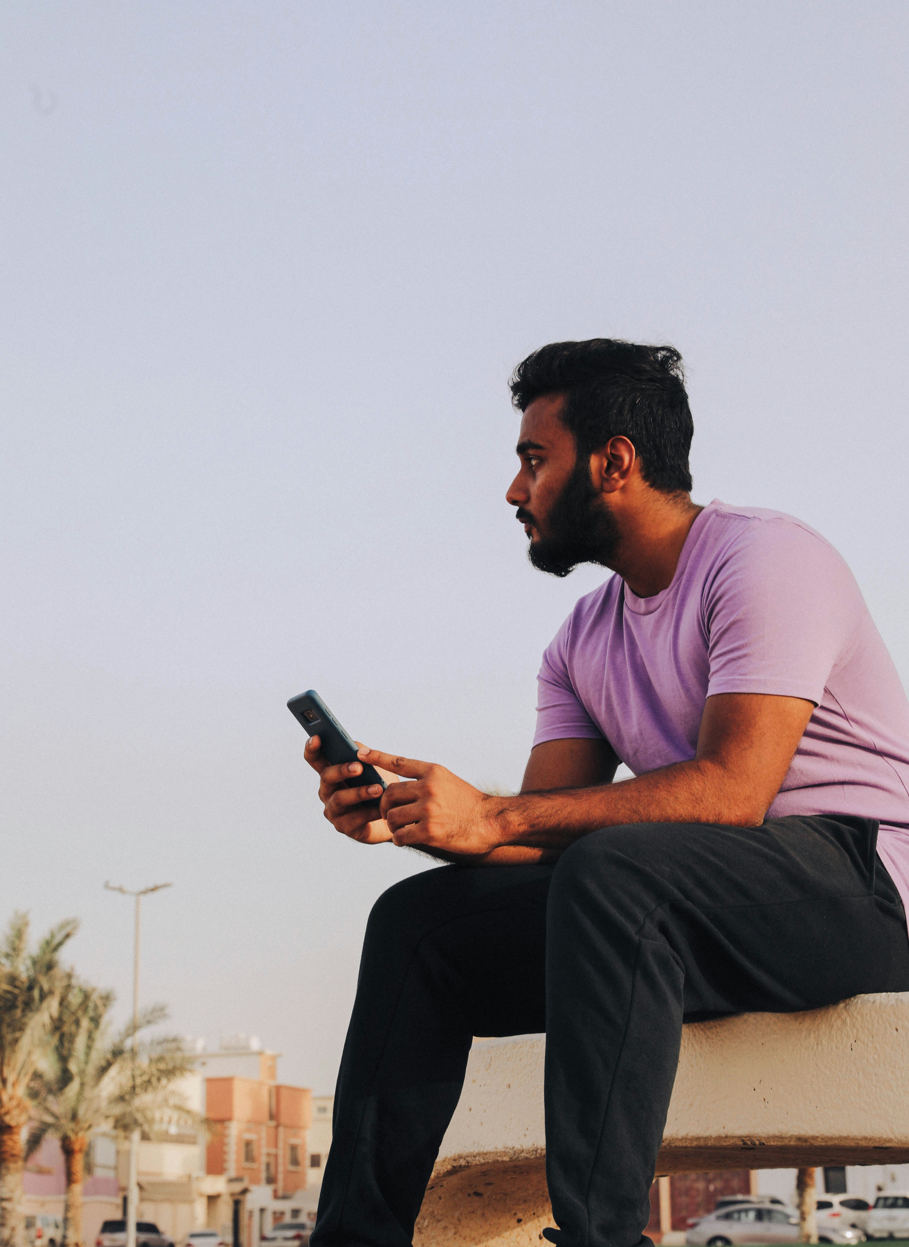 Man sitting on a ledge, absorbed in his phone against a backdrop of urban architecture and palm trees.