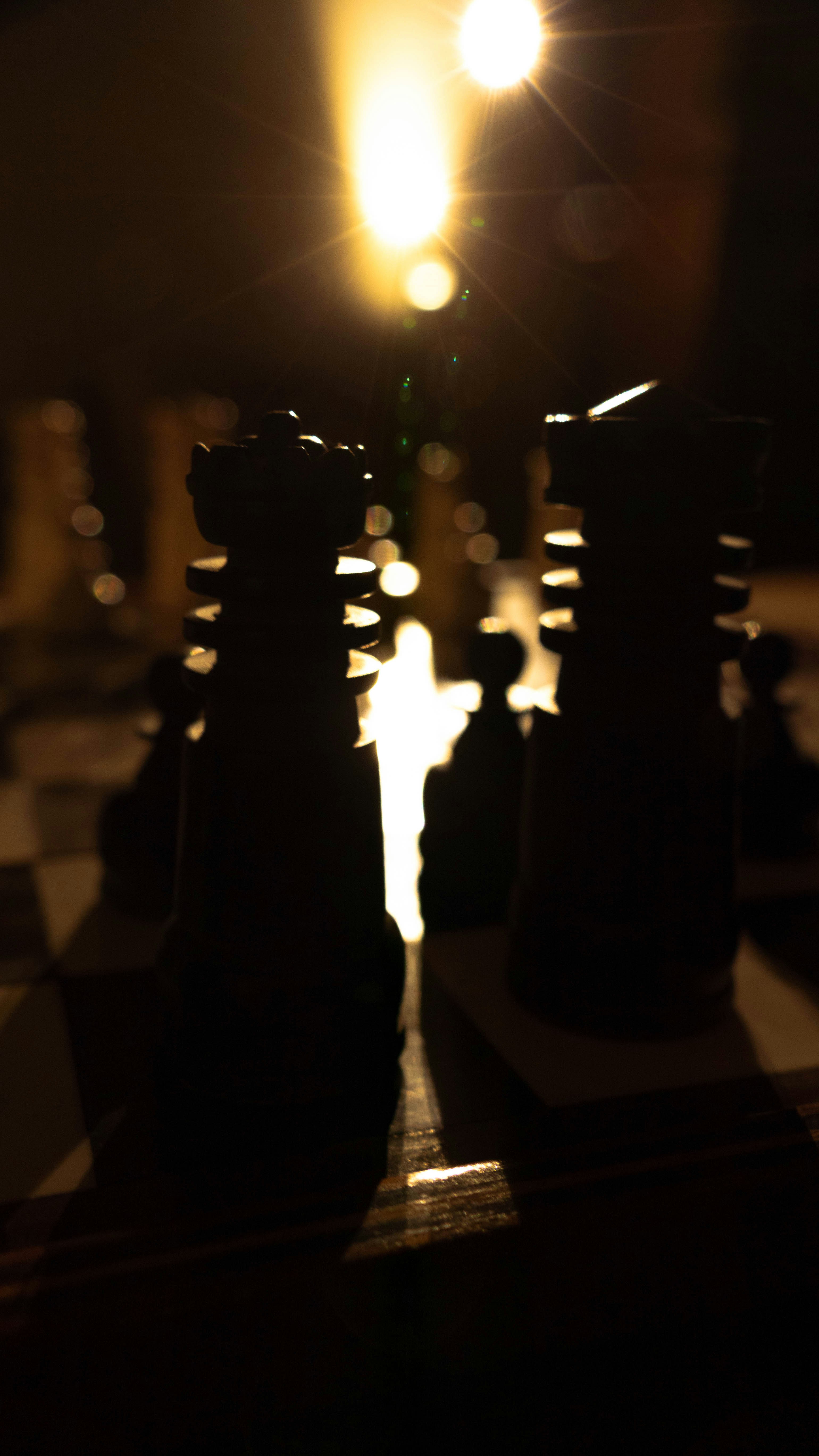 Silhouetted chess pieces stand in a dimly lit setting, with a bright light source creating an ethereal glow in the background.