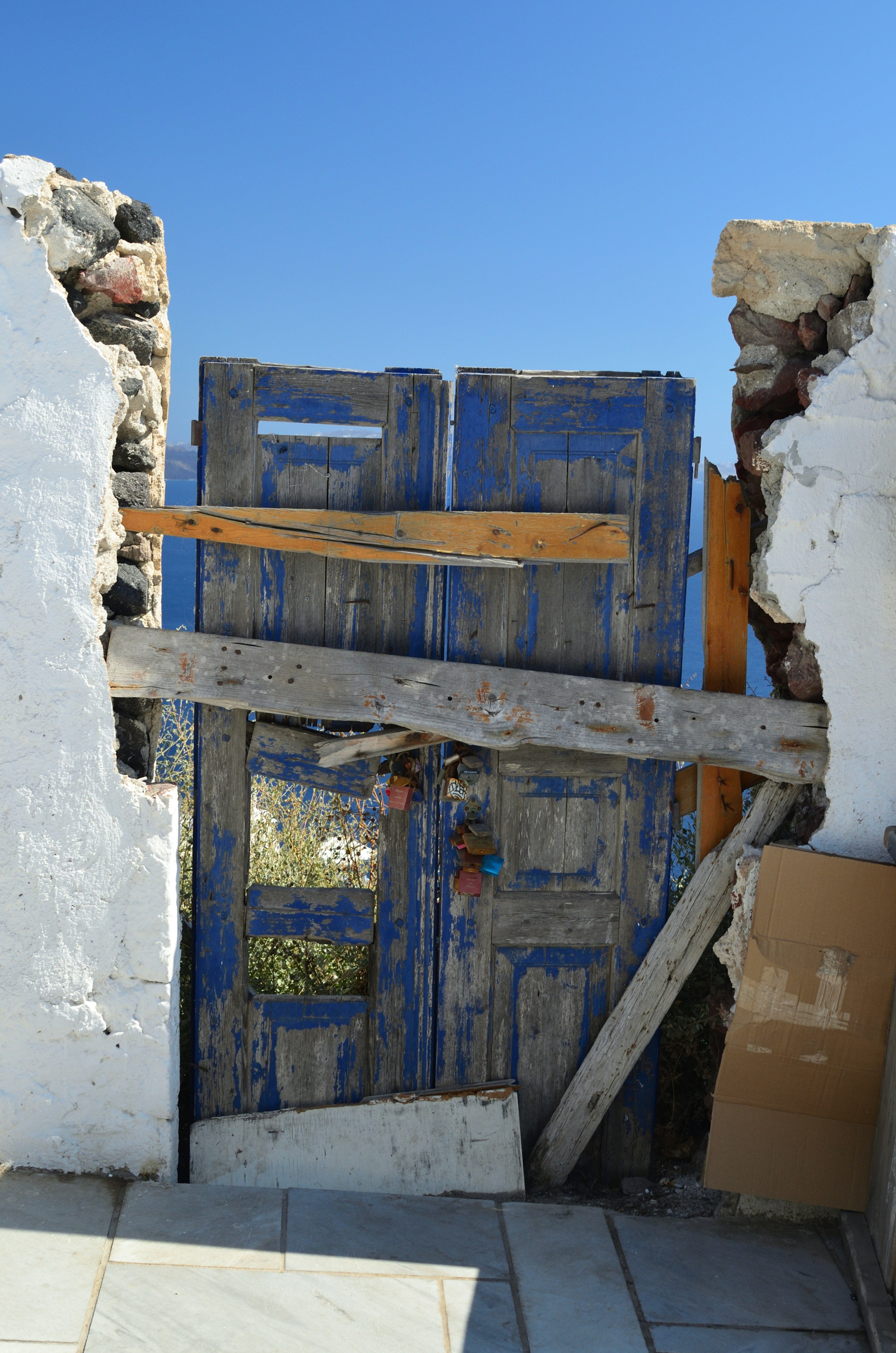 Weathered blue doors secured with wooden beams, framed by crumbling white walls, hint at a once vibrant entrance overlooking the sea.