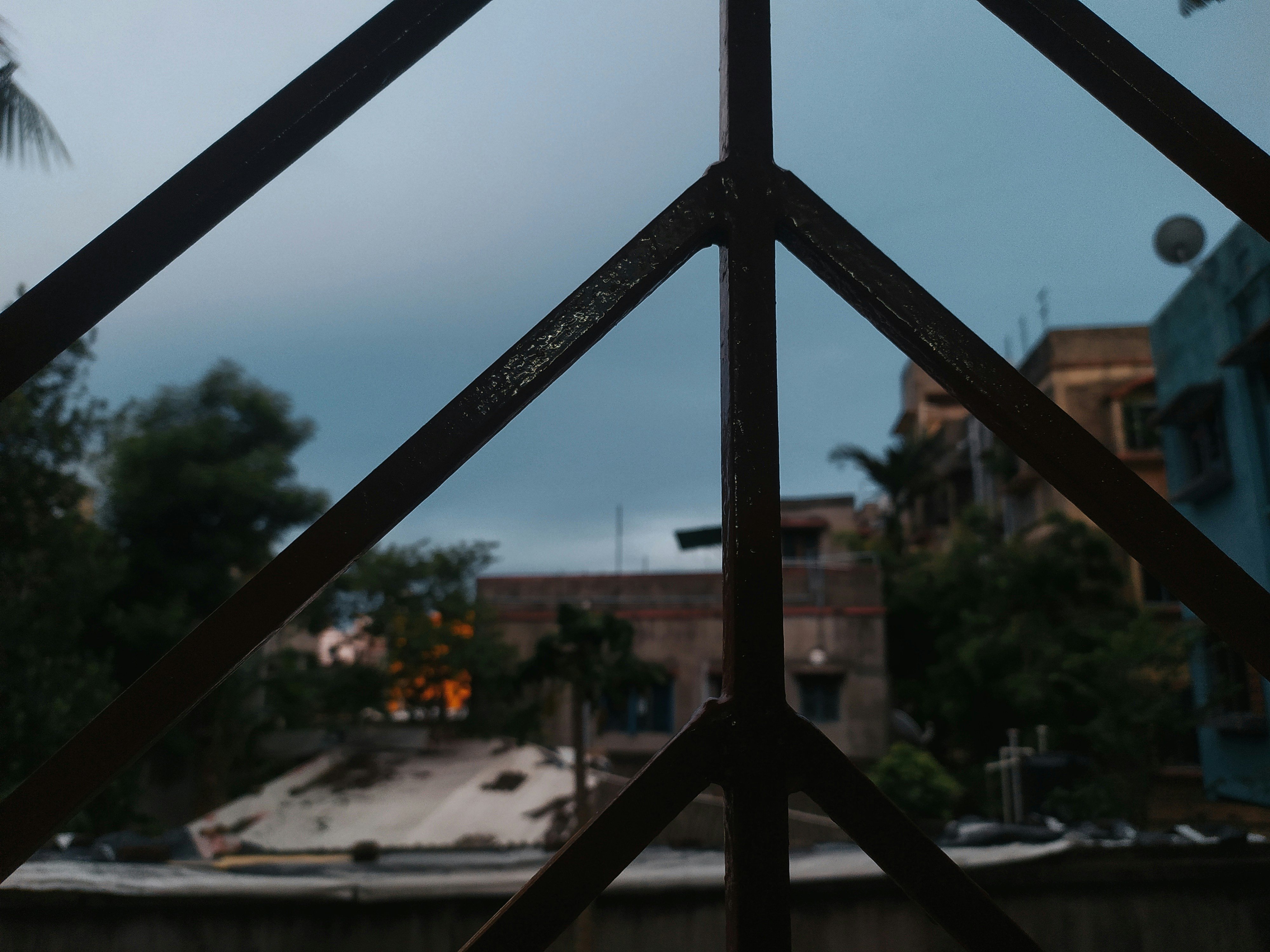 Urban scene framed by a dark metal lattice, with soft morning light revealing distant buildings and trees beyond the gate.