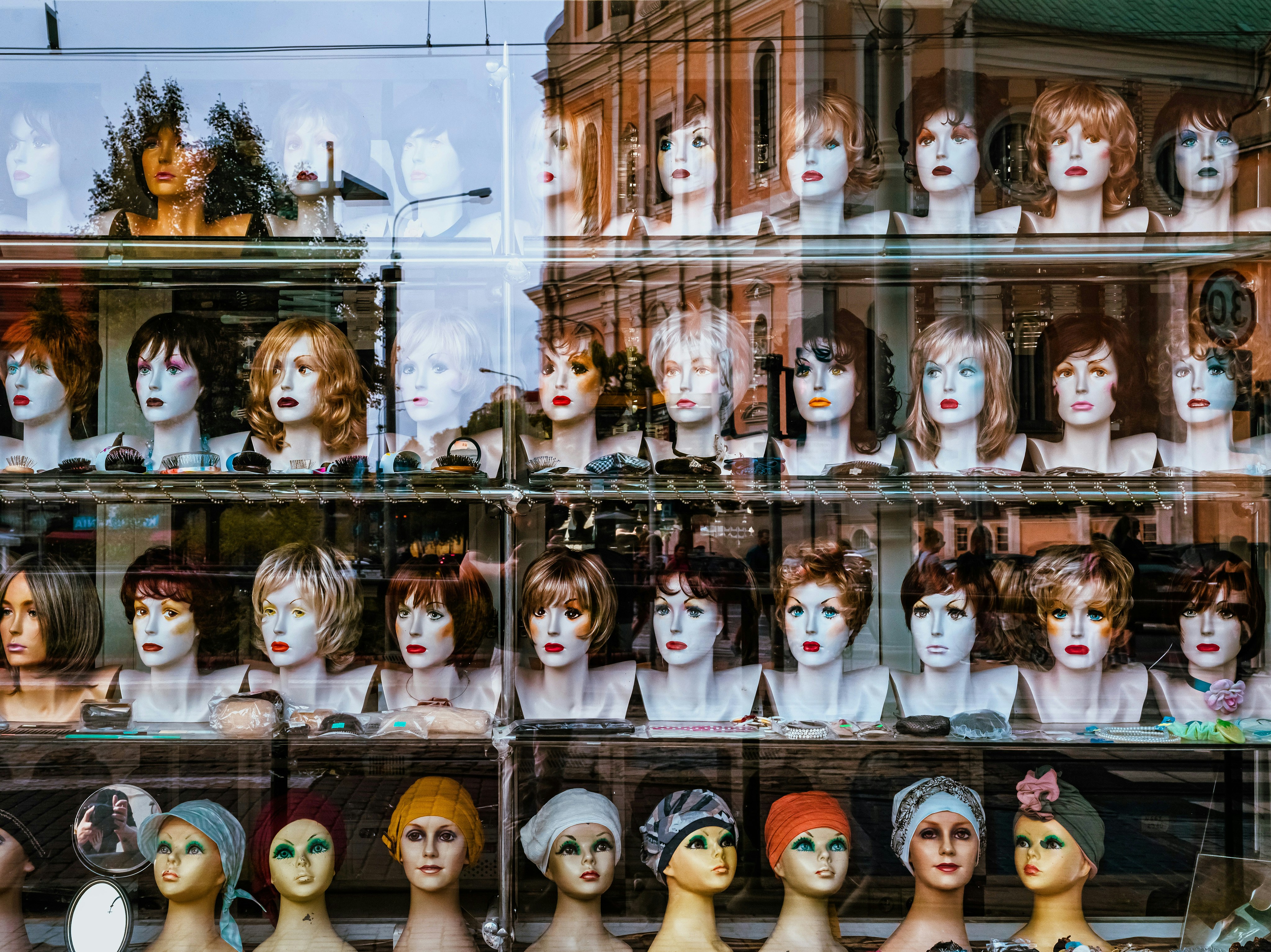 Storefront window displaying a grid of mannequin heads wearing wigs, with reflections of a building in the glass.