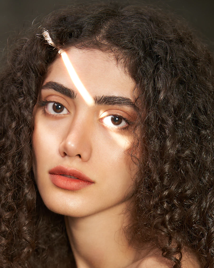 Close-up portrait of a woman with curly dark hair, strong brows, and a band of sunlight across her forehead and eye. Neutral background keeps attention on her face.