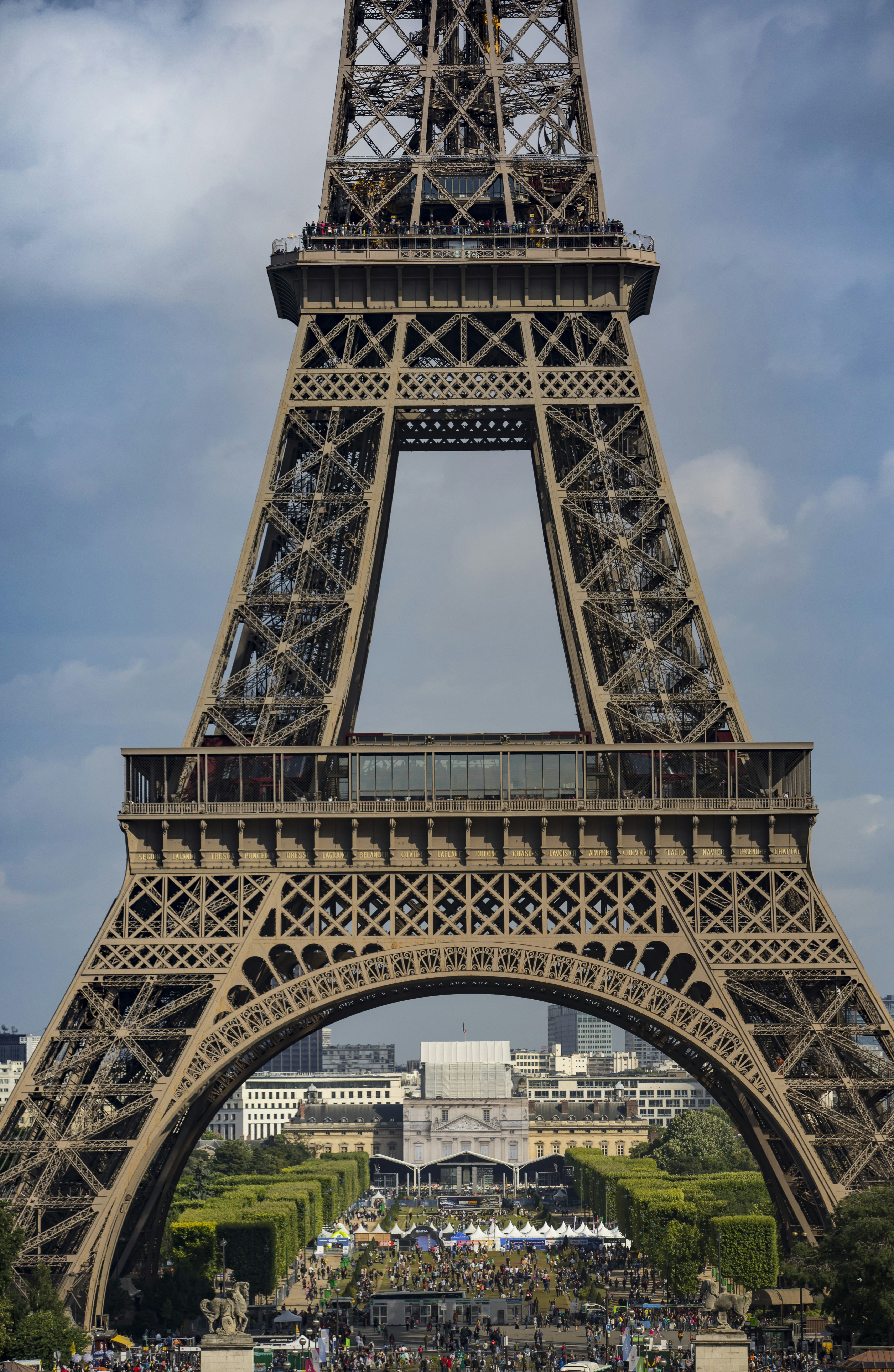 Eiffel Tower viewed from beneath, showcasing its intricate iron lattice work against a cloudy sky. The bustling gardens below add life to the scene.