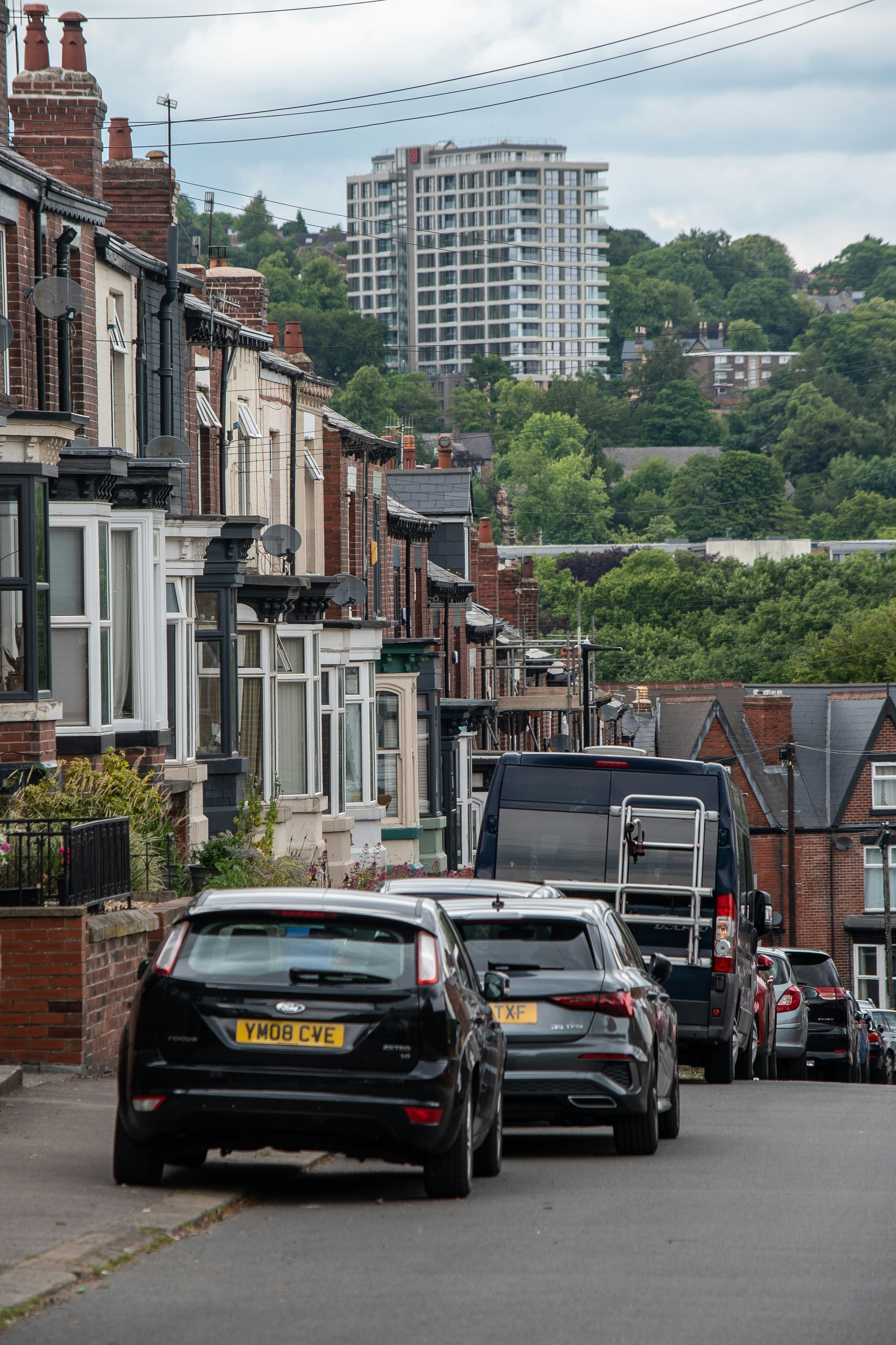 Residential street lined with traditional houses and parked cars, leading to a backdrop of modern high-rise buildings and lush greenery.