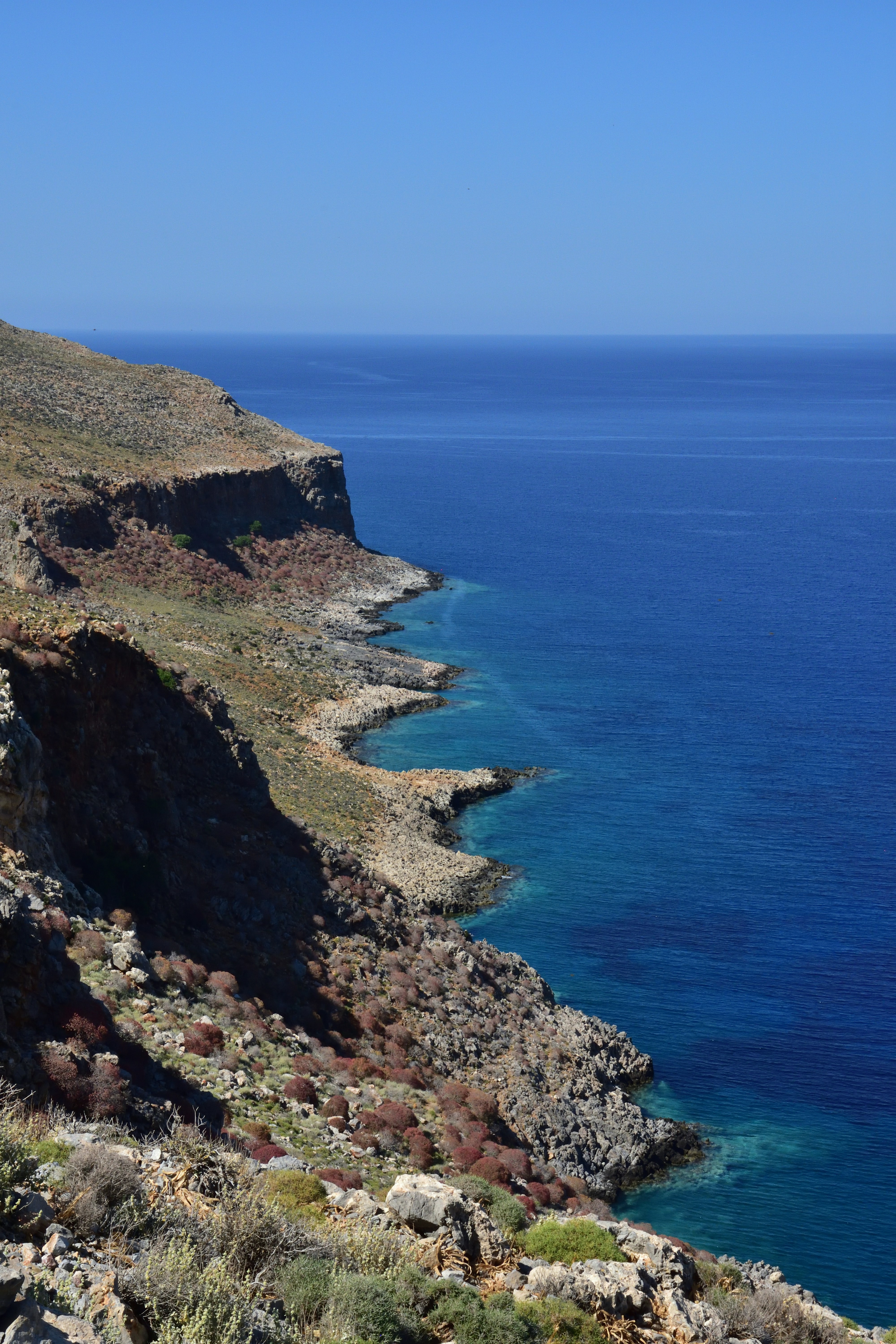 Rocky coastline meeting tranquil blue waters under a clear sky. The rugged terrain showcases the natural beauty of the landscape.