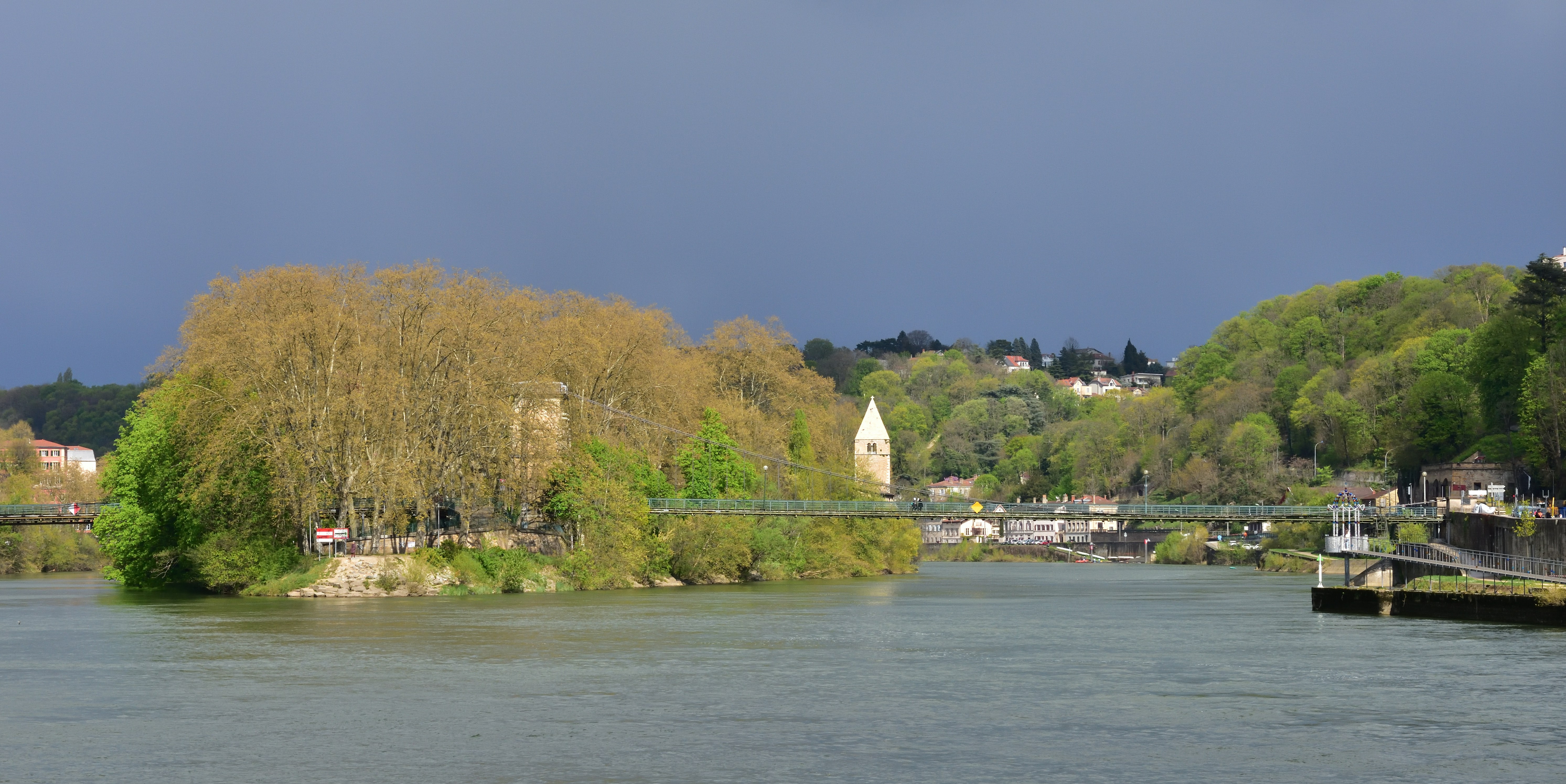 Lush green island surrounded by a river, with a distant tower peeking through the trees under an ominous sky.