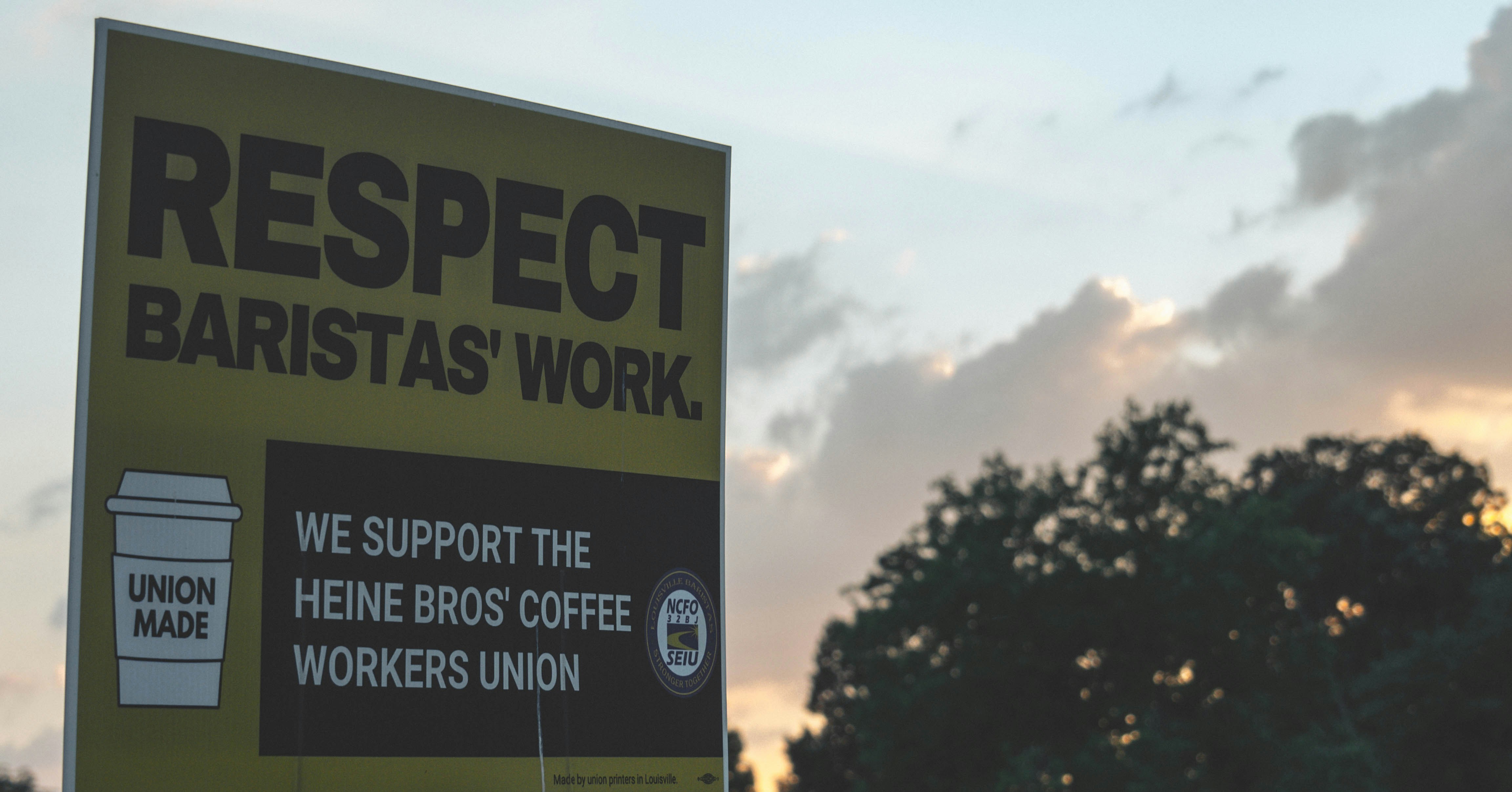 Protest sign supporting coffee workers against a backdrop of silhouetted trees at sunset.