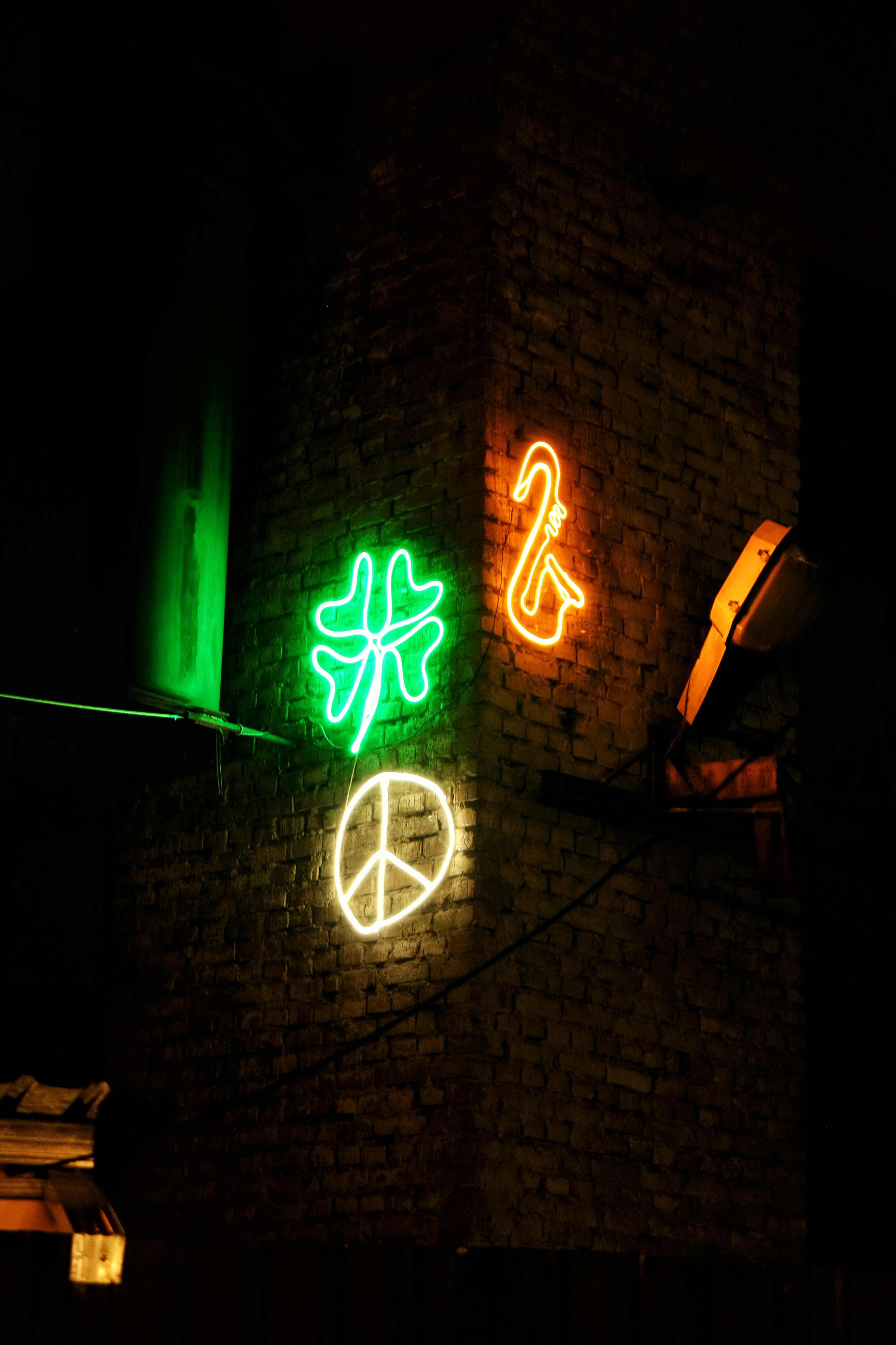 Neon Glyphs on a Dark Brick Wall: Green Clover, Peace Sign, and an Orange Curve