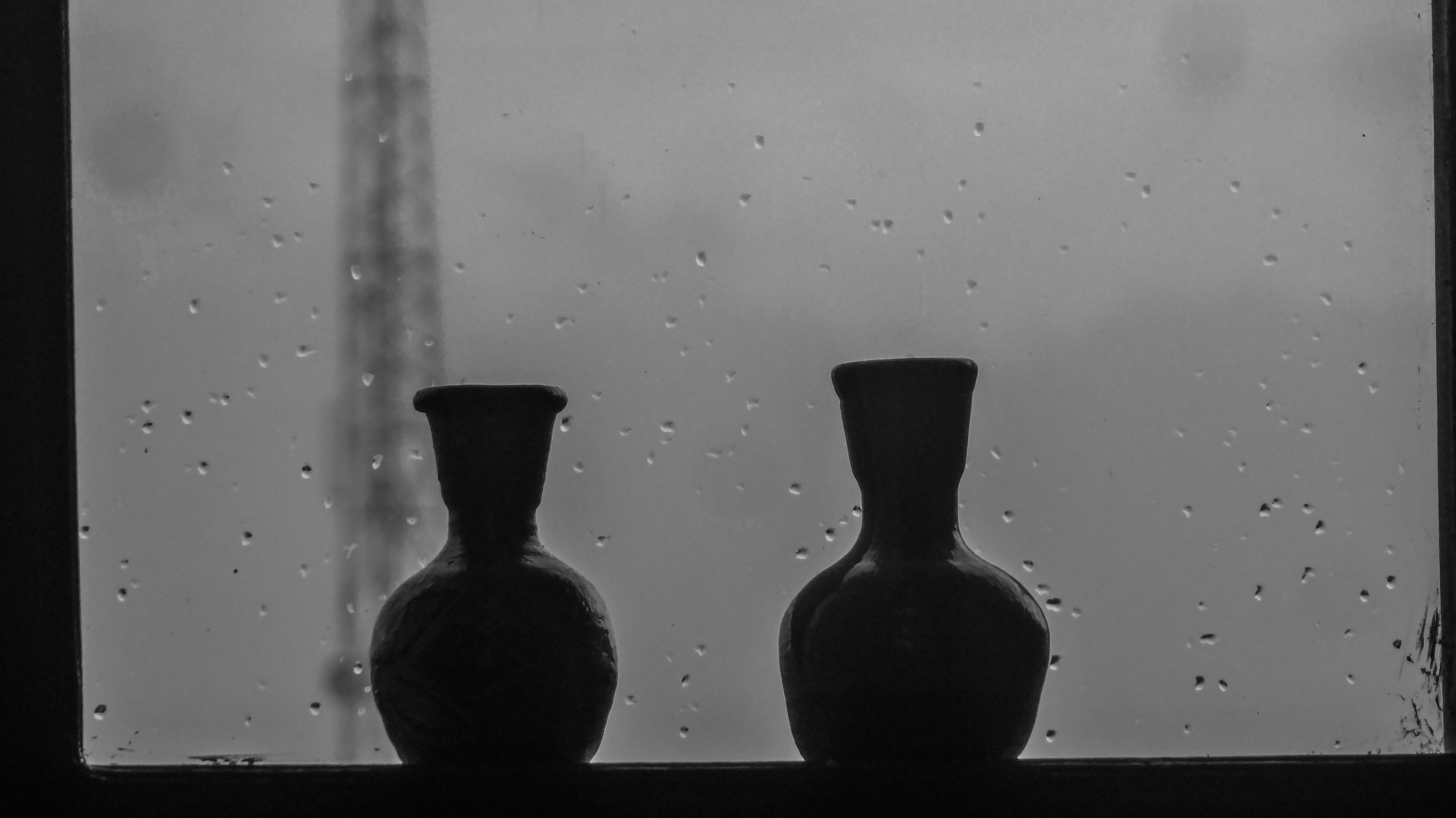 Two vases silhouetted against a rainy window, creating a moody atmosphere. The droplets enhance the sense of tranquility.
