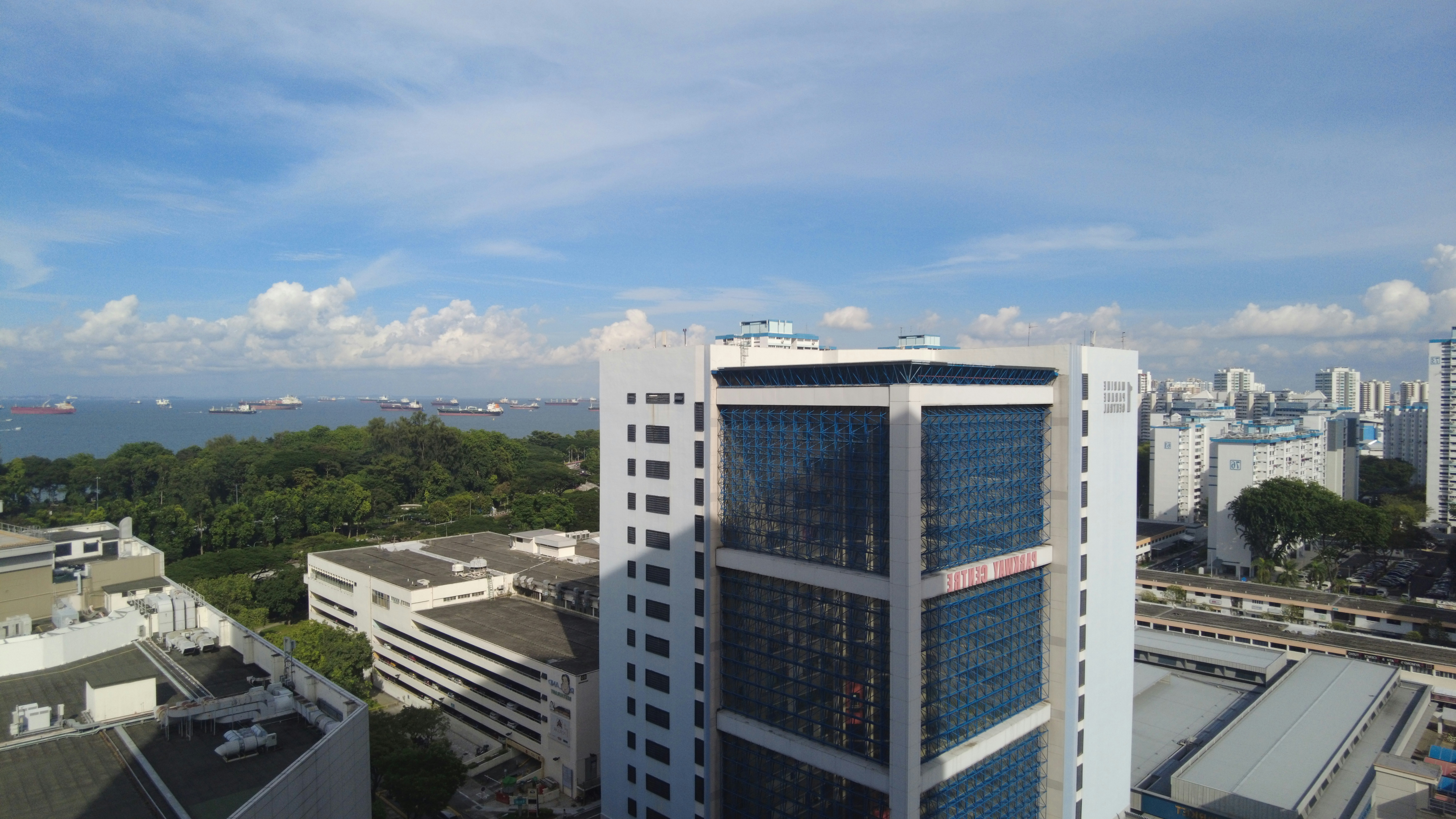 Photograph of a coastal cityscape dominated by a white, glass-fronted high-rise, with a green park and the sea visible beyond. Ships dot the horizon, completing the harbor backdrop.