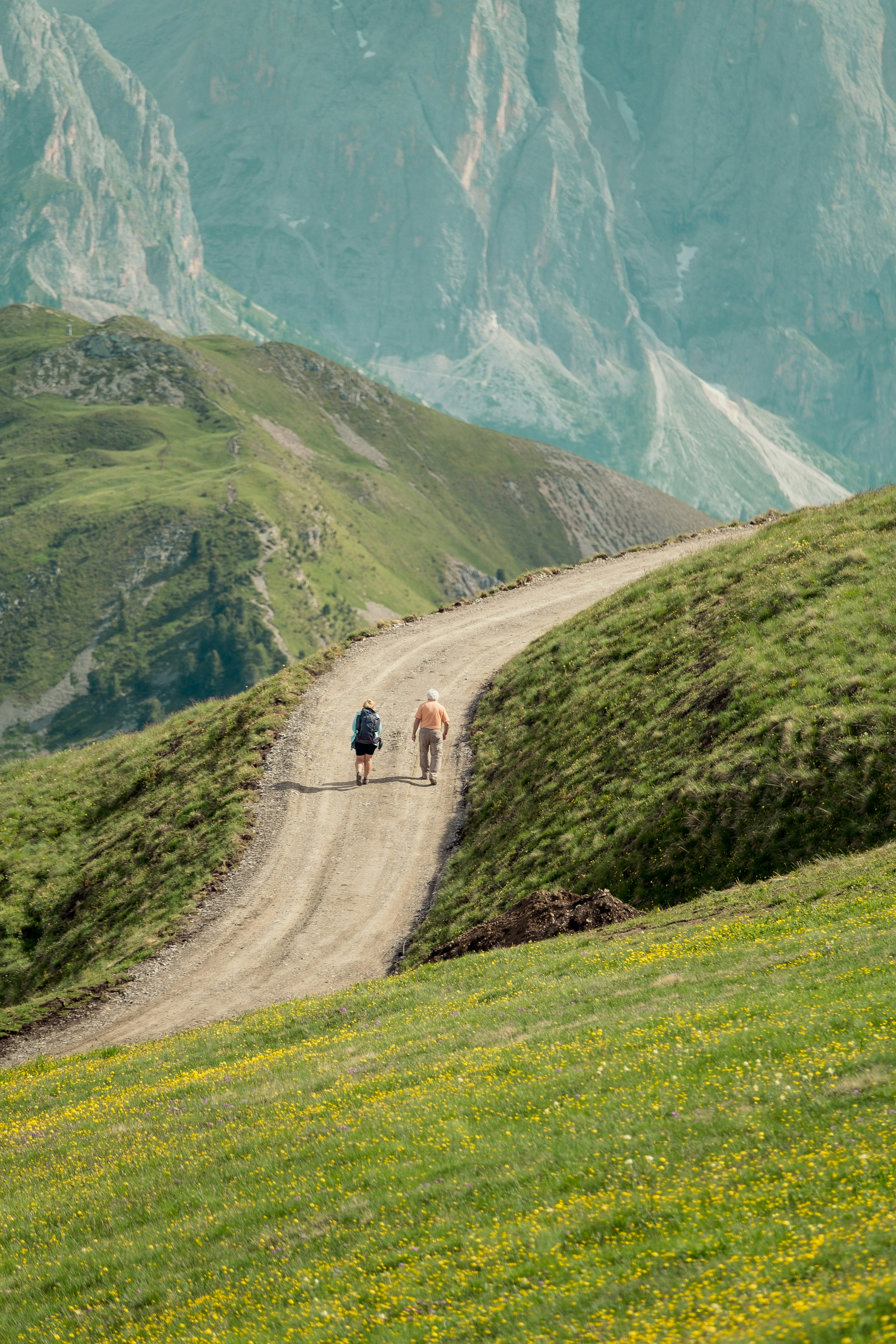 Two hikers on a winding ridge beneath towering alpine cliffs