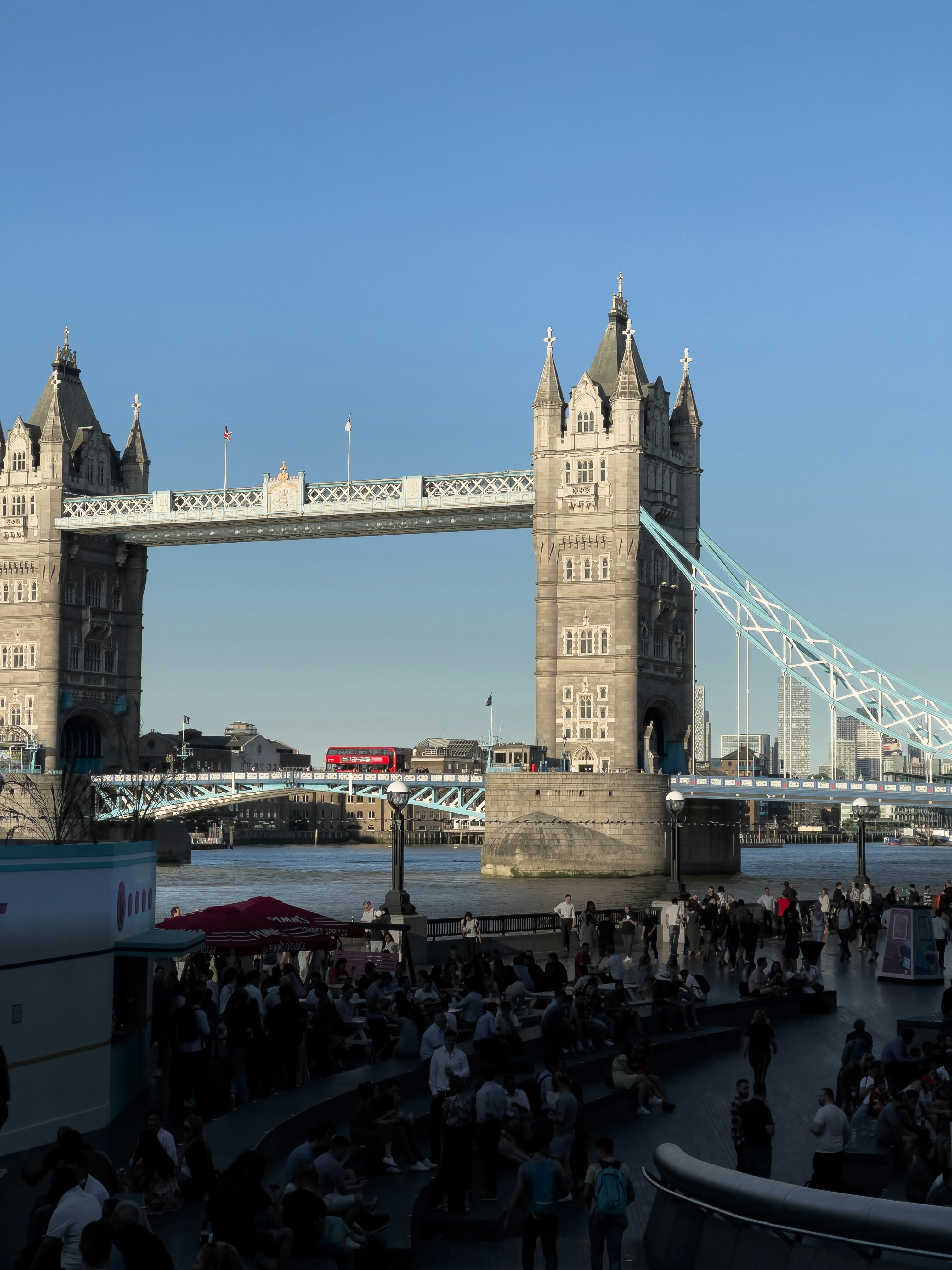 Tower Bridge stands majestically over the Thames, framed by a vibrant blue sky, with crowds enjoying the riverside promenade.