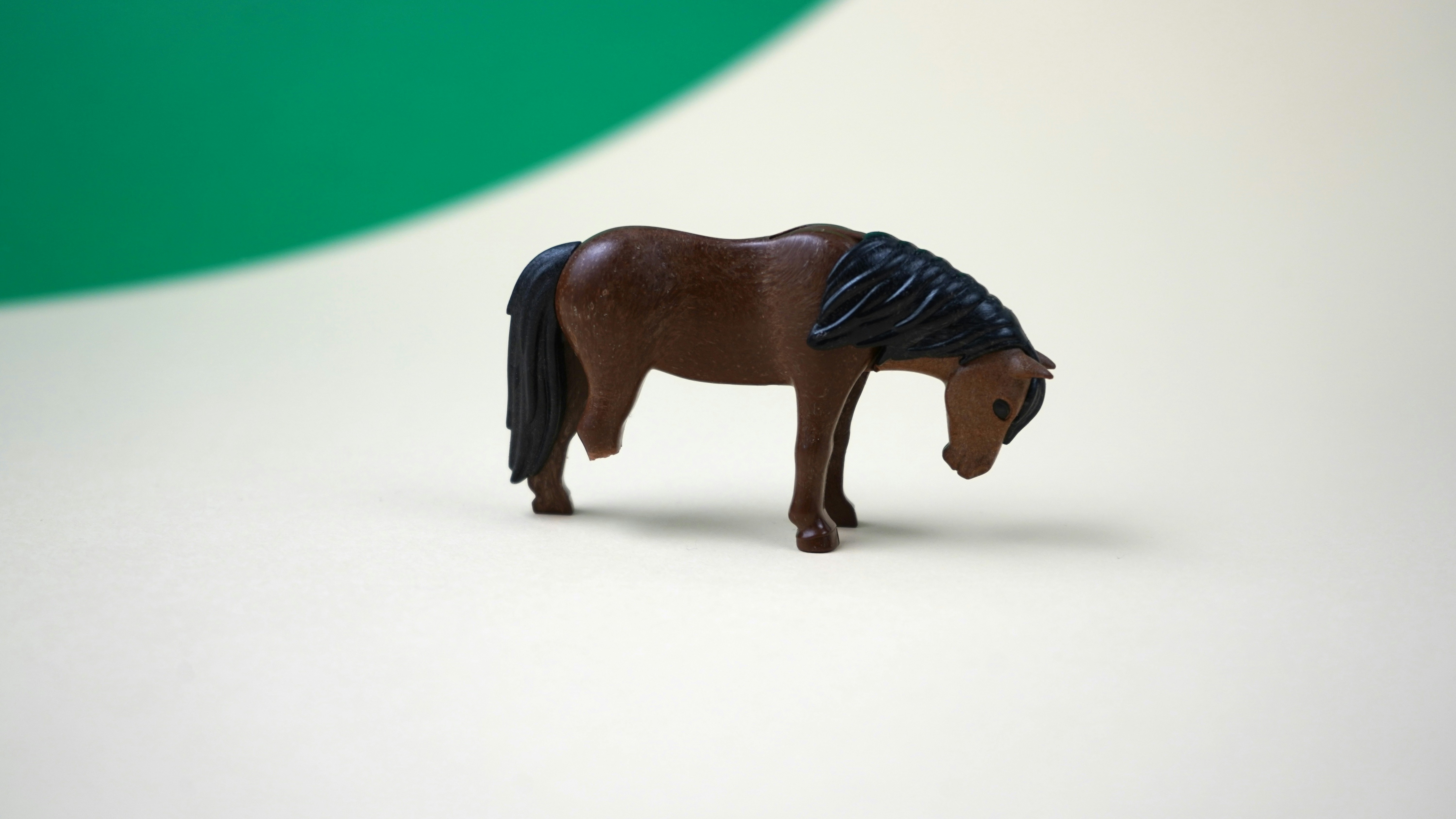 A toy horse with a glossy finish stands on a soft surface, its head lowered as if in deep thought. The background features a smooth gradient of green and cream.