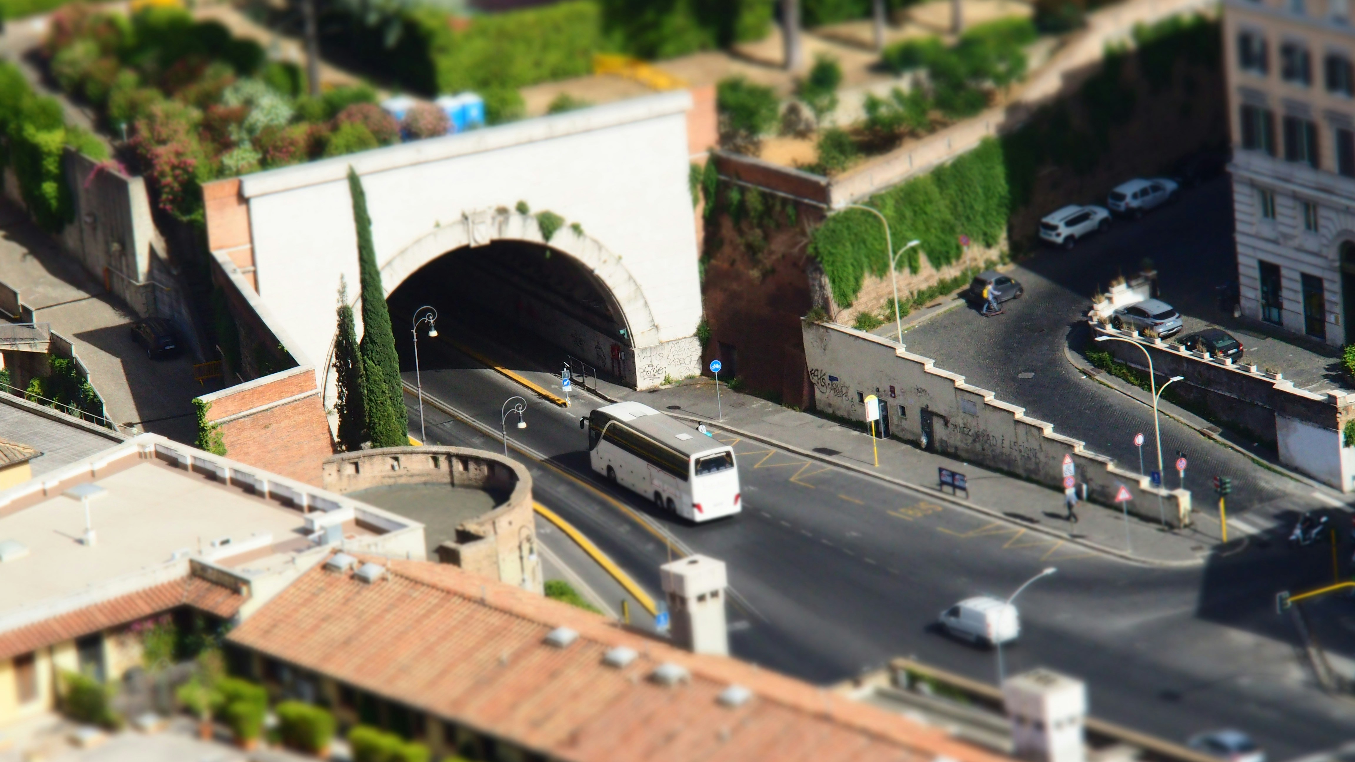 A bus approaches a tunnel entrance in a bustling urban setting, surrounded by greenery and architectural details.