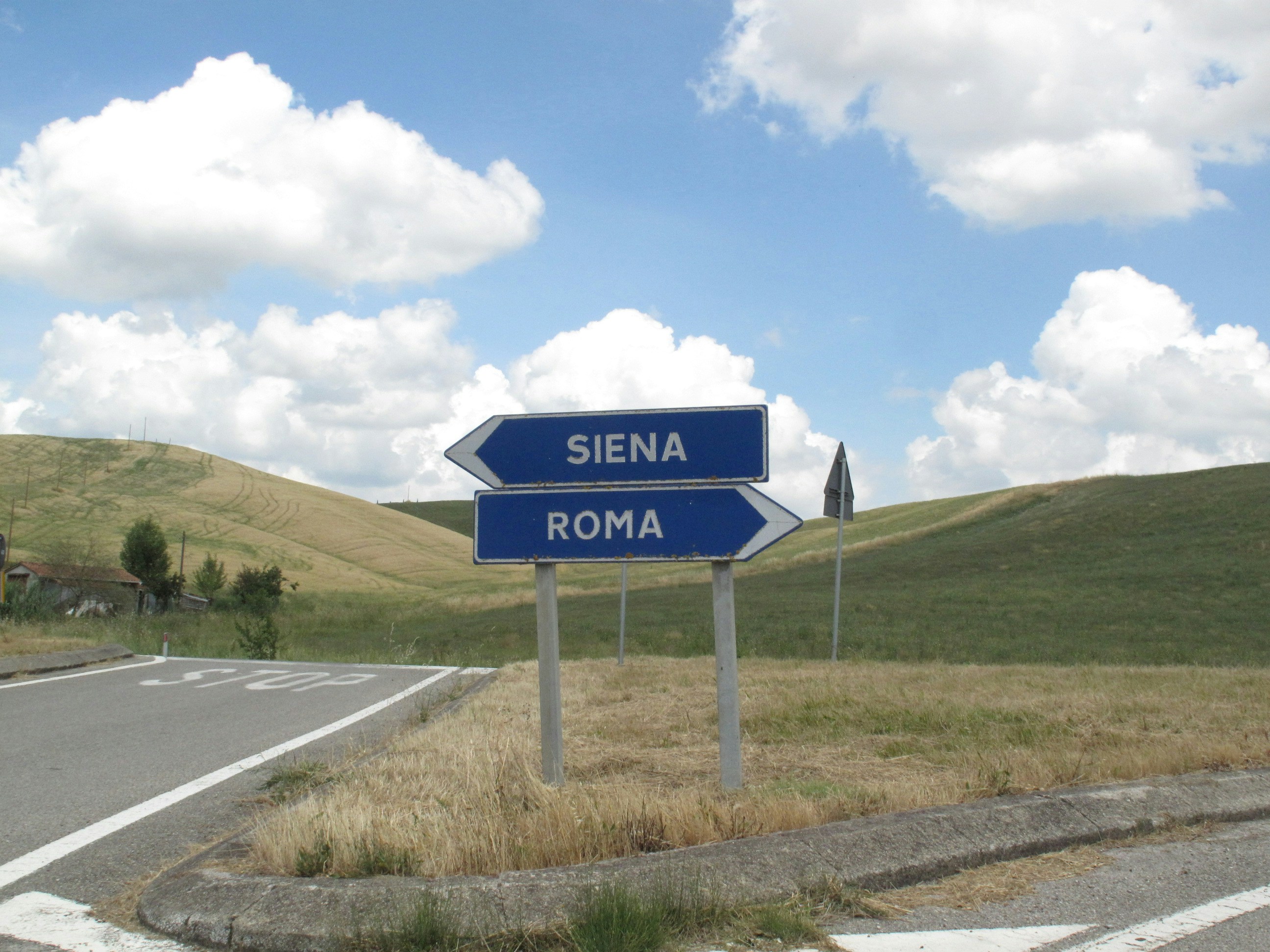 Directional road signs pointing towards Siena and Roma against a backdrop of rolling hills and fluffy clouds.