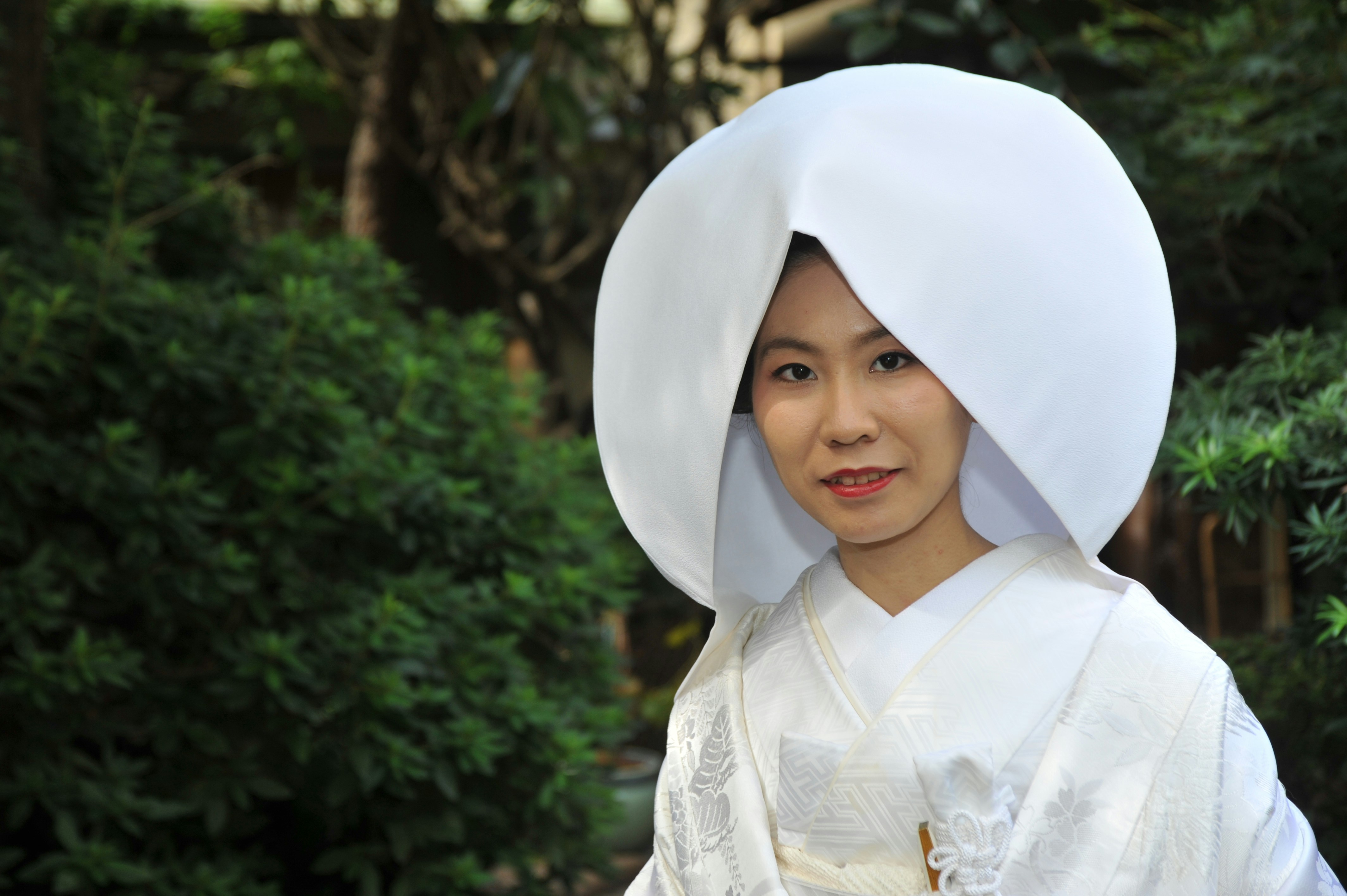Woman dressed in traditional Japanese wedding attire, featuring a distinctive white headpiece, set against a lush green backdrop.
