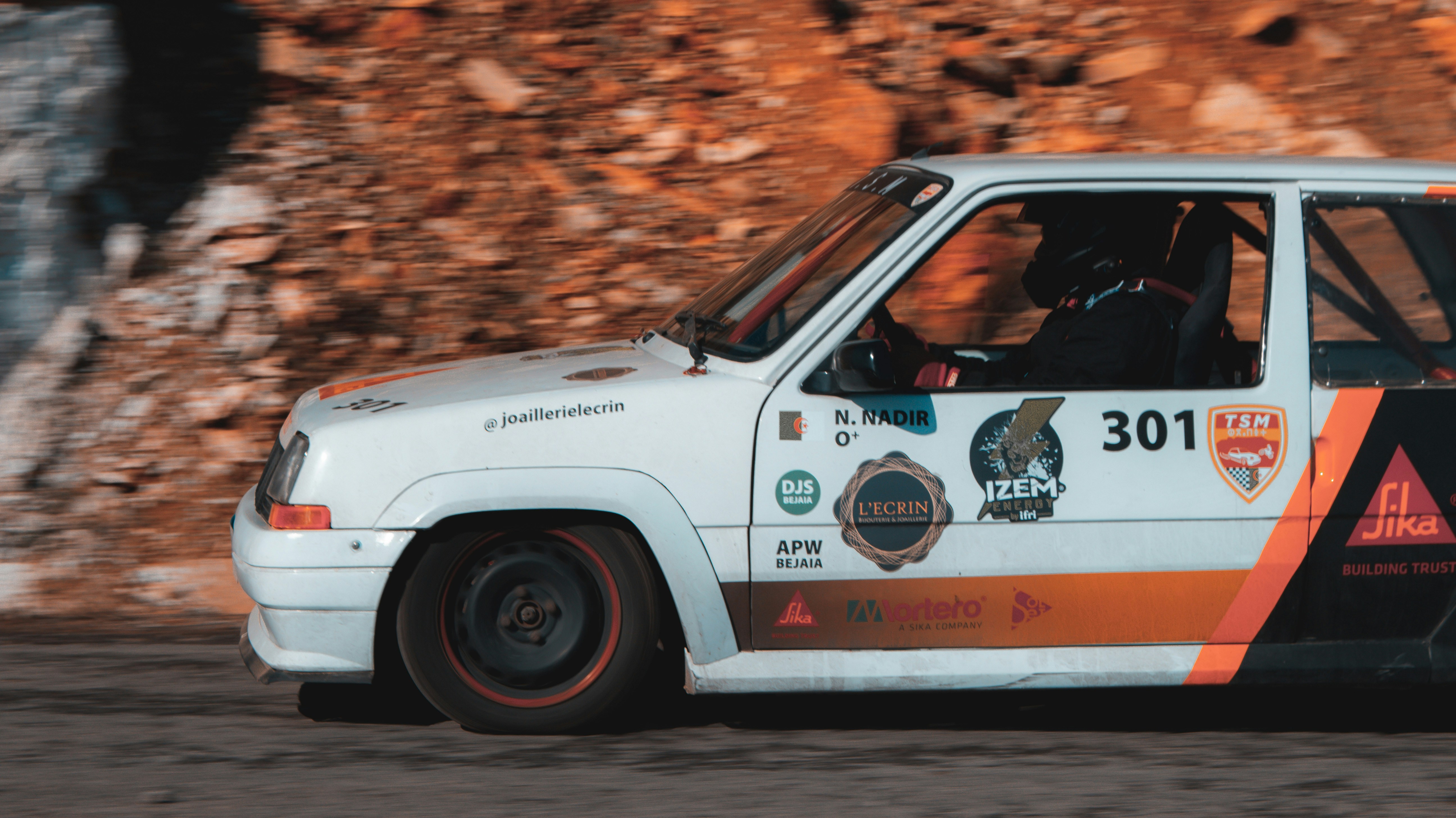 White rally car speeding on a track with a rocky backdrop.