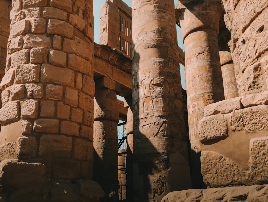 Massive weathered sandstone columns carved with hieroglyphs inside an ancient temple ruin, lit by warm sunlight and framed by dark shadows.