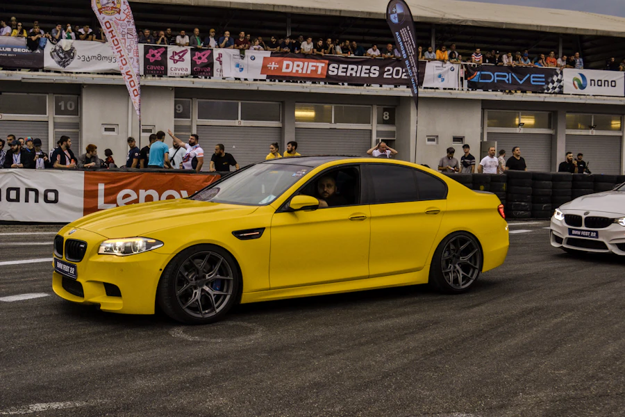 Yellow BMW sedan moving along a racetrack with the driver visible through the window and a crowd on the pit stands behind it.
