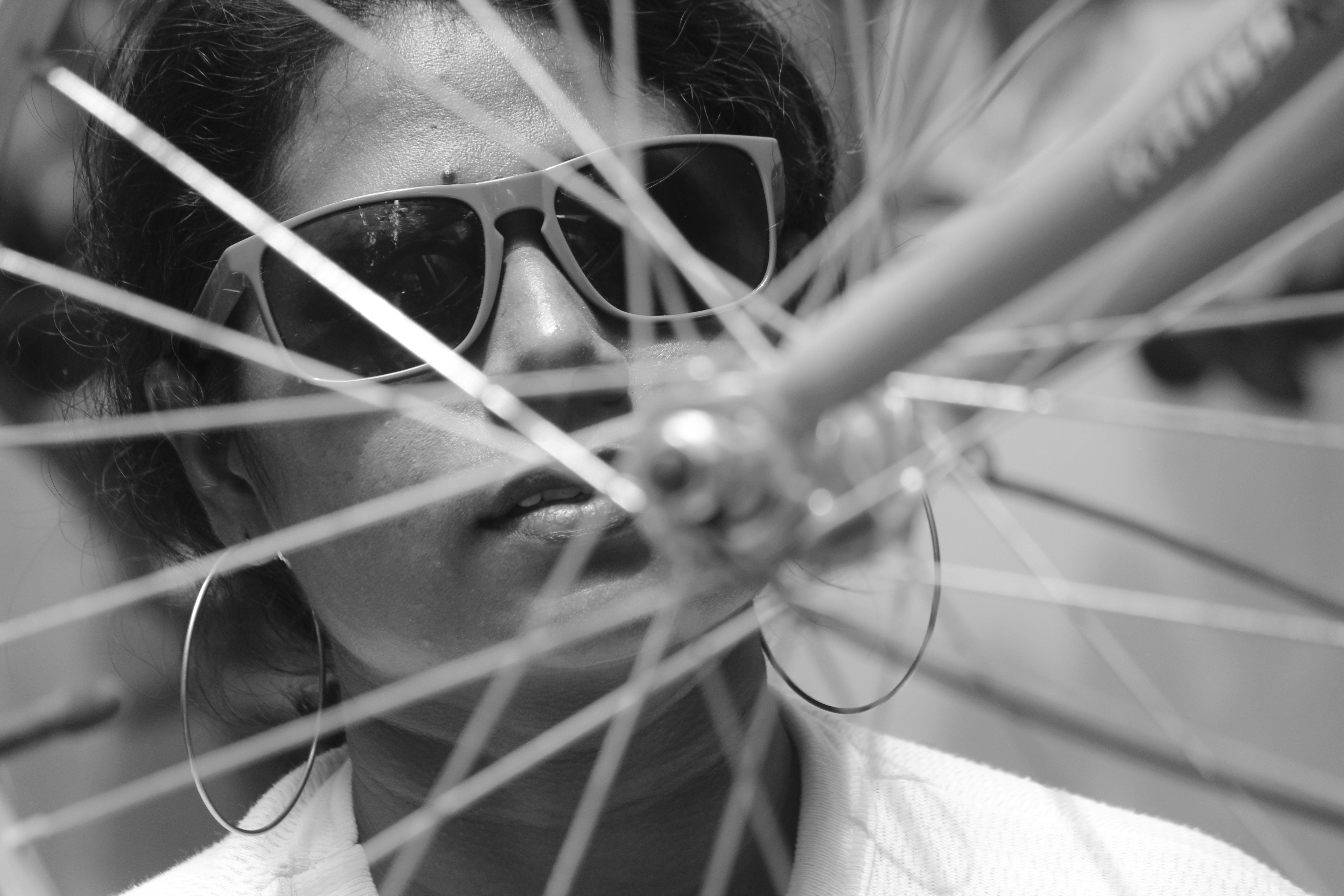 Monochrome portrait of a person wearing sunglasses, partially obscured by bicycle spokes; a bold, graphic composition that centers on the subject's face peeking through the spoke pattern.