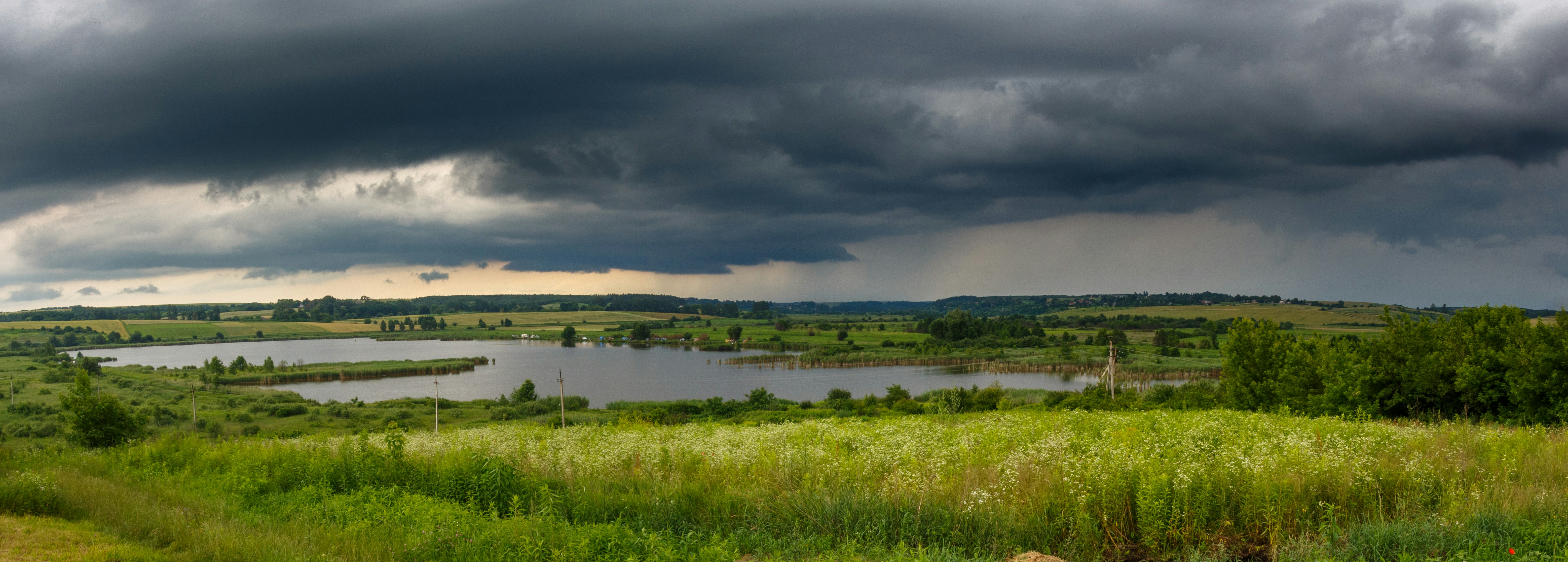 A panoramic view of a serene lake surrounded by lush greenery under an ominous sky filled with dark clouds. The scene evokes a sense of tranquility mixed with anticipation.