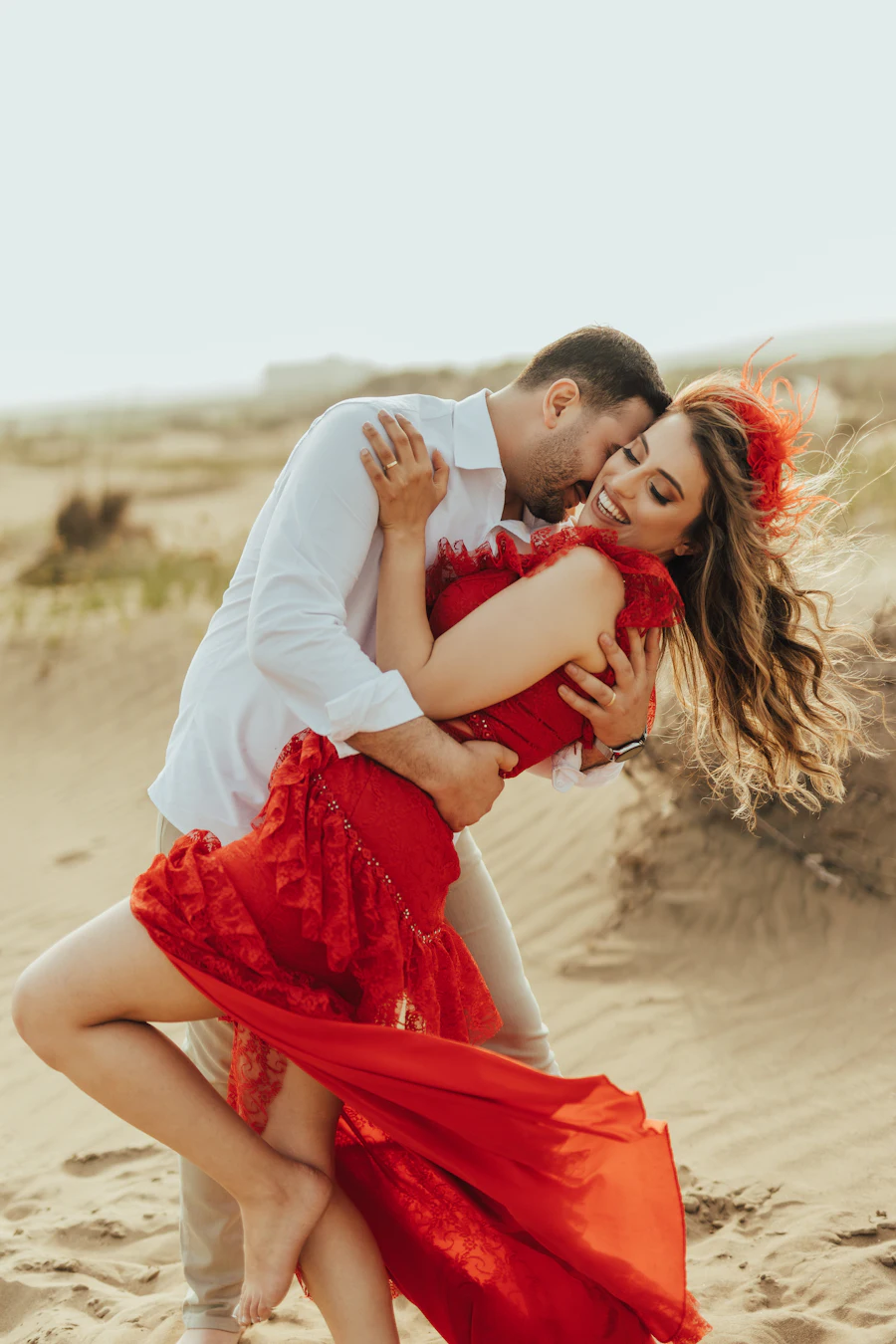 A couple embraces closely in a sandy desert landscape, with the woman in a bright red dress and the man in a white shirt. Wind lifts her hair and dress as they lean into a tender kiss.