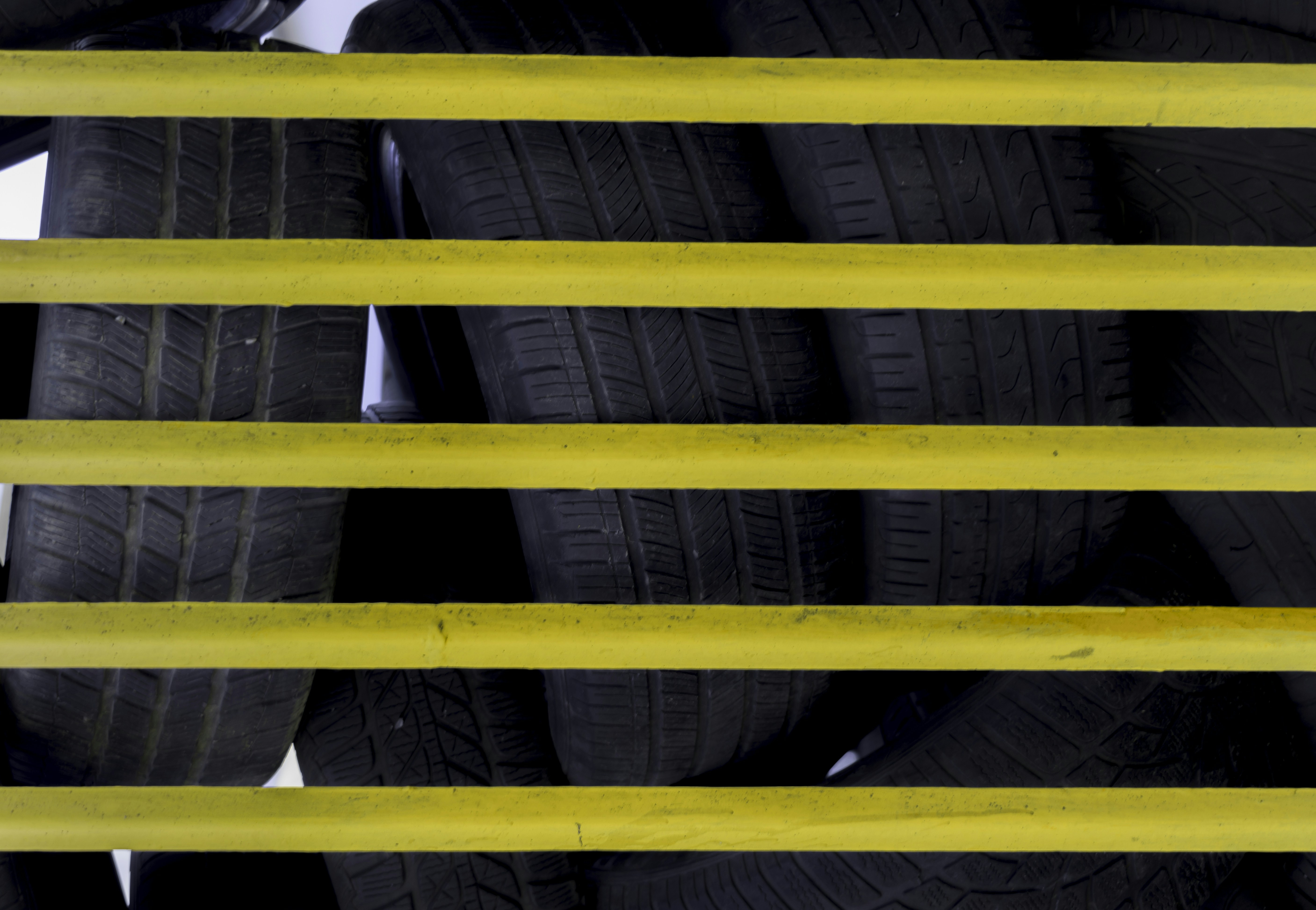 Photograph of stacked black tires viewed through evenly spaced yellow bars, creating a striped foreground. The dense rubber textures contrast with the bright barrier lines.