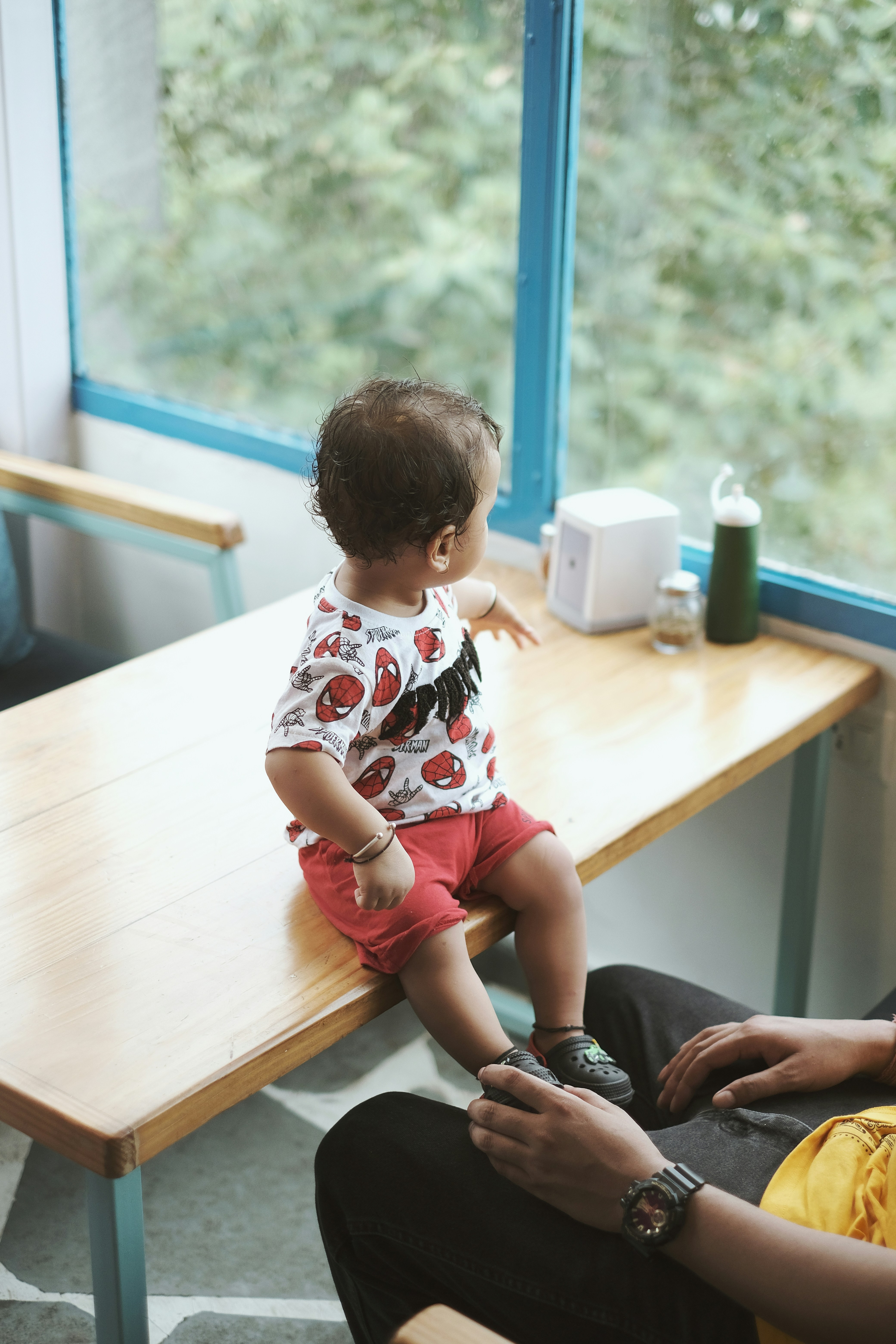 Window-side Wonder: A toddler perched on a wooden desk surveys the greenery beyond