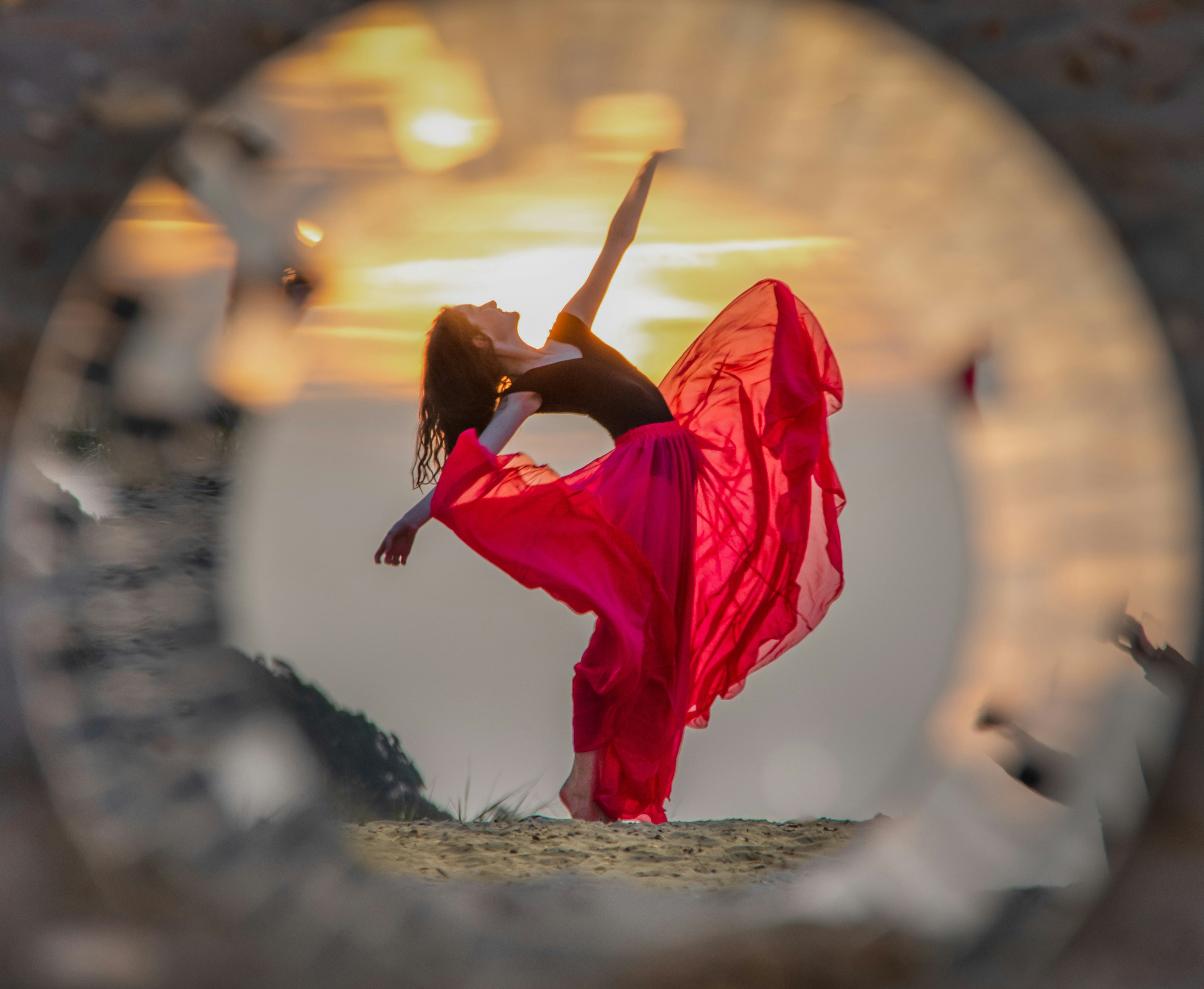 A dancer in a flowing red skirt gracefully poses against a vibrant sunset, framed through a circular opening. The scene captures the interplay of movement and light.