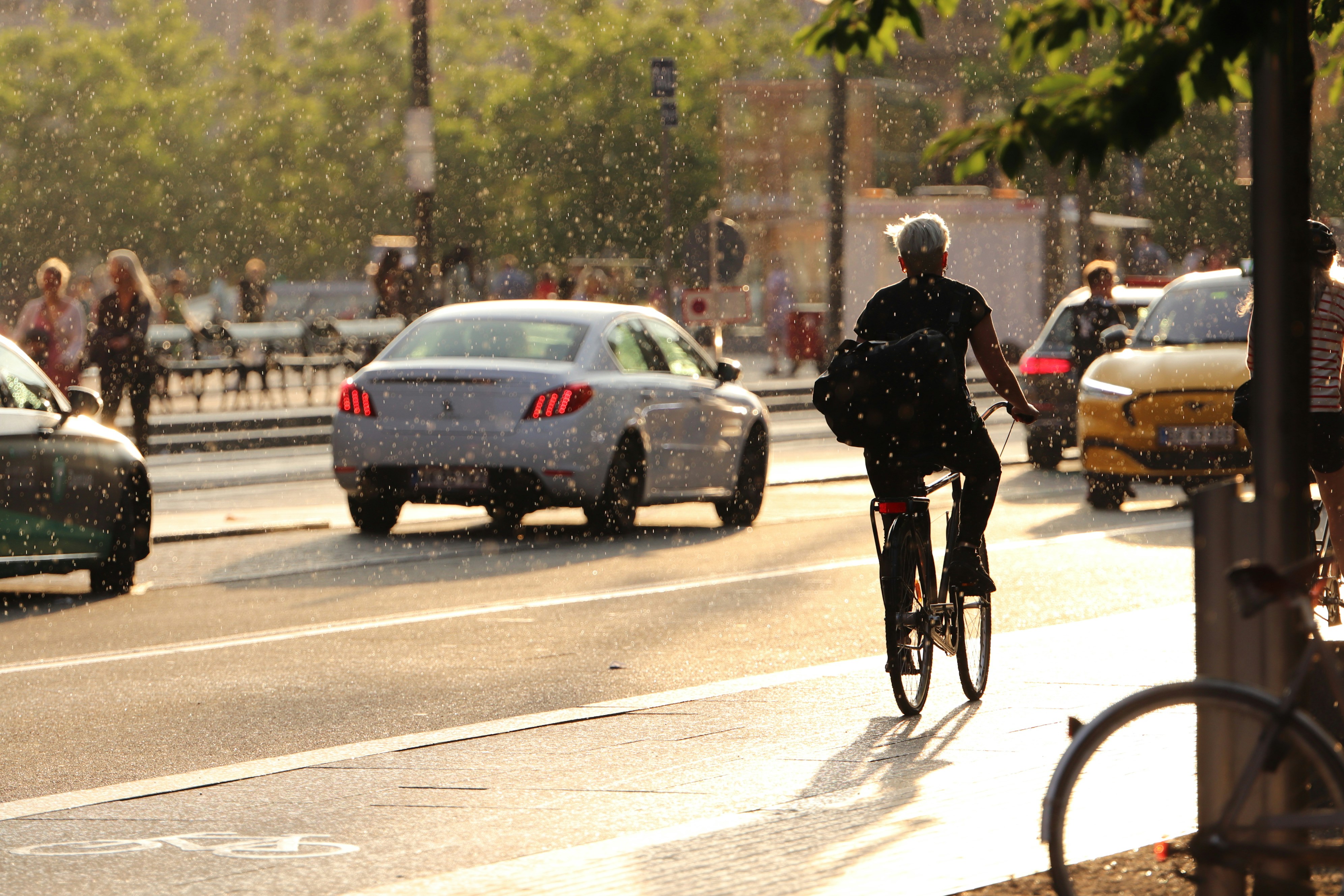 A cyclist navigates through a busy street as sunlight filters through raindrops, creating a vibrant urban scene. The atmosphere is alive with movement and reflections.