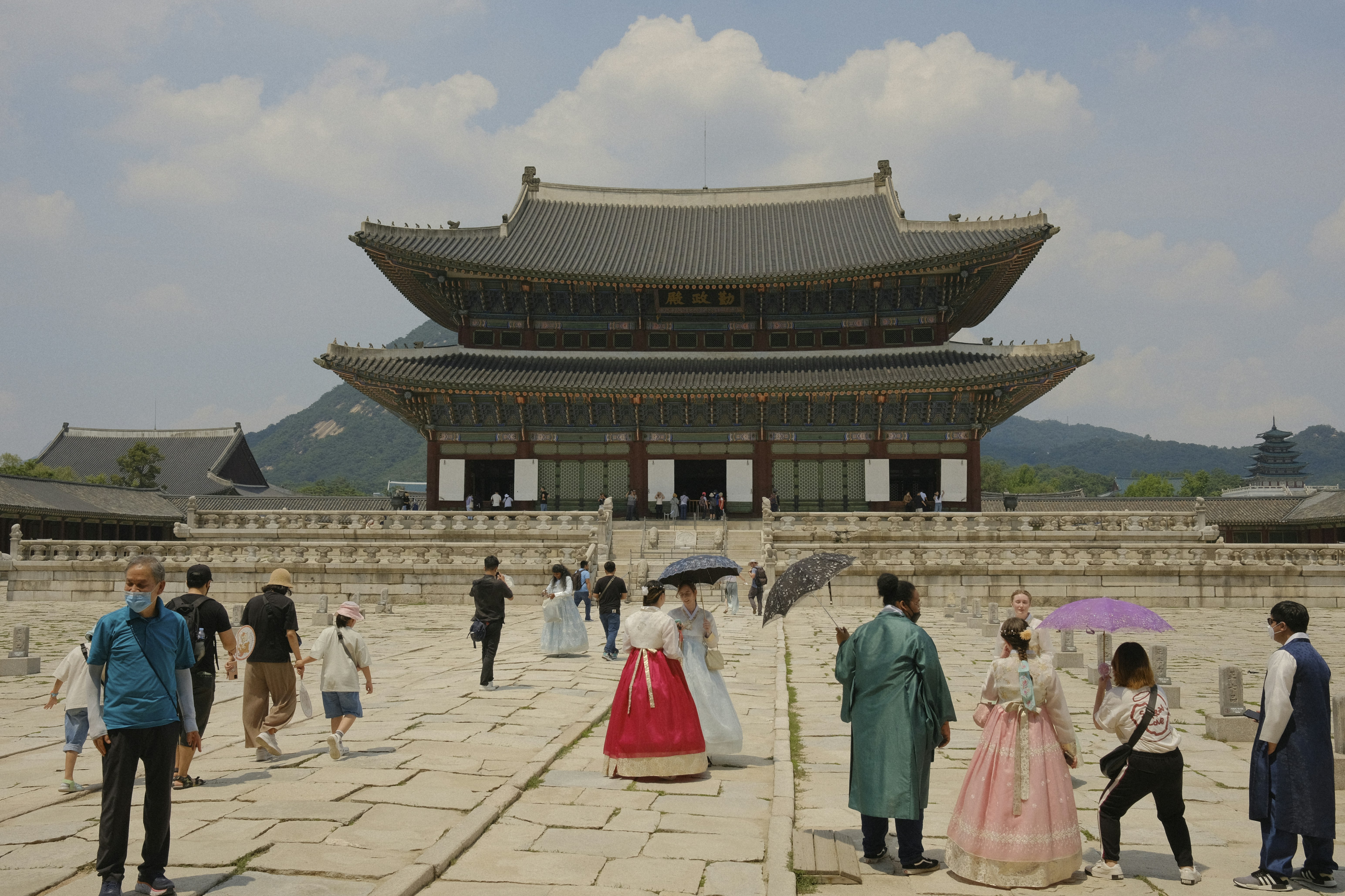 This captivating image showcases a traditional palace with its grand architectural design, set against a backdrop of soft blue skies and fluffy clouds. Visitors in colorful traditional attire and casual modern clothing create a vibrant mix of past and present, walking along the expansive stone courtyard. The image's warm tones and natural lighting highlight the intricate details of the palace, creating a harmonious and inviting atmosphere.