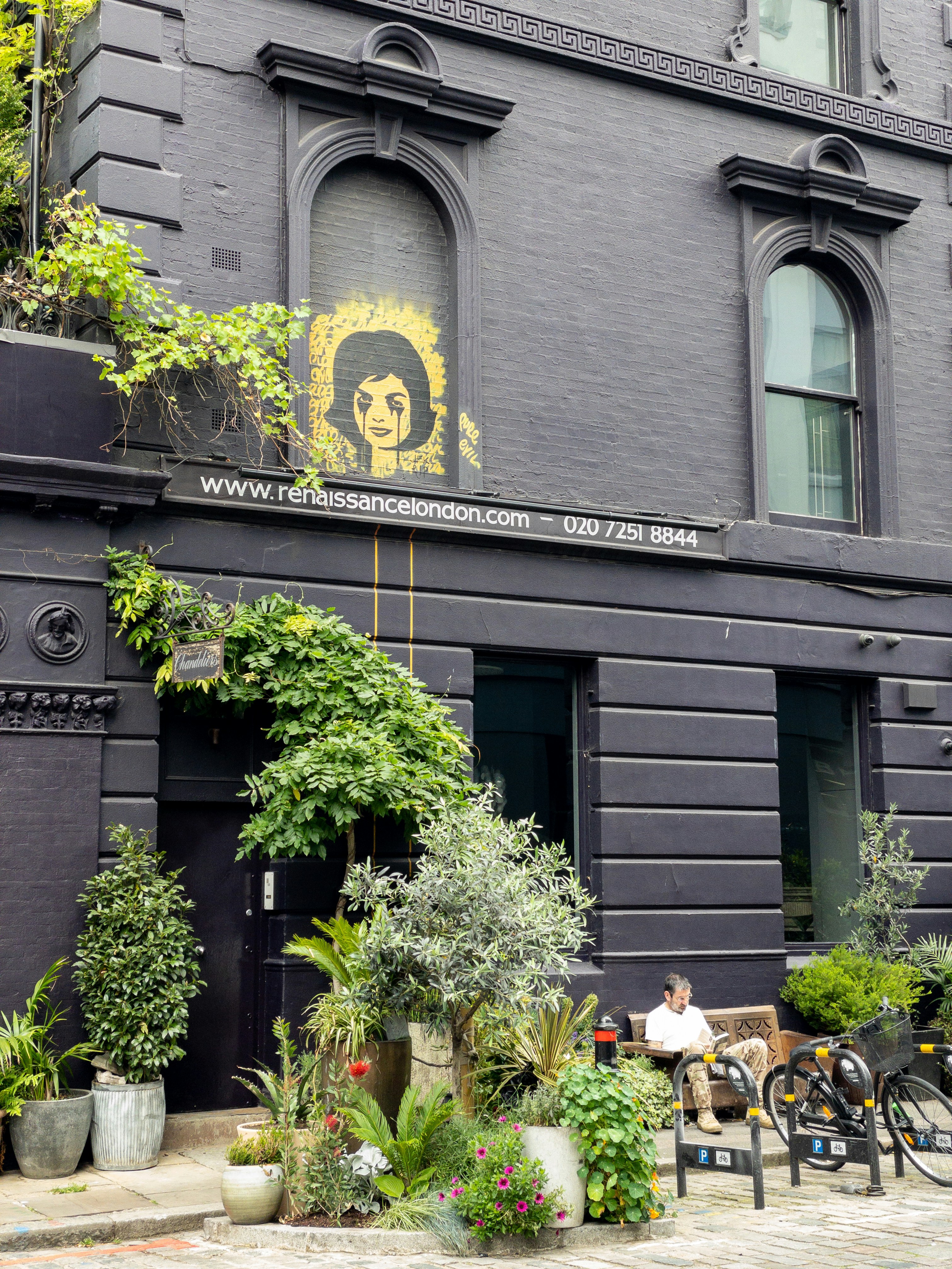 A vibrant mural of a woman framed by lush greenery on a dark building facade, with a person relaxing in the foreground. The scene combines urban art and natural elements seamlessly.