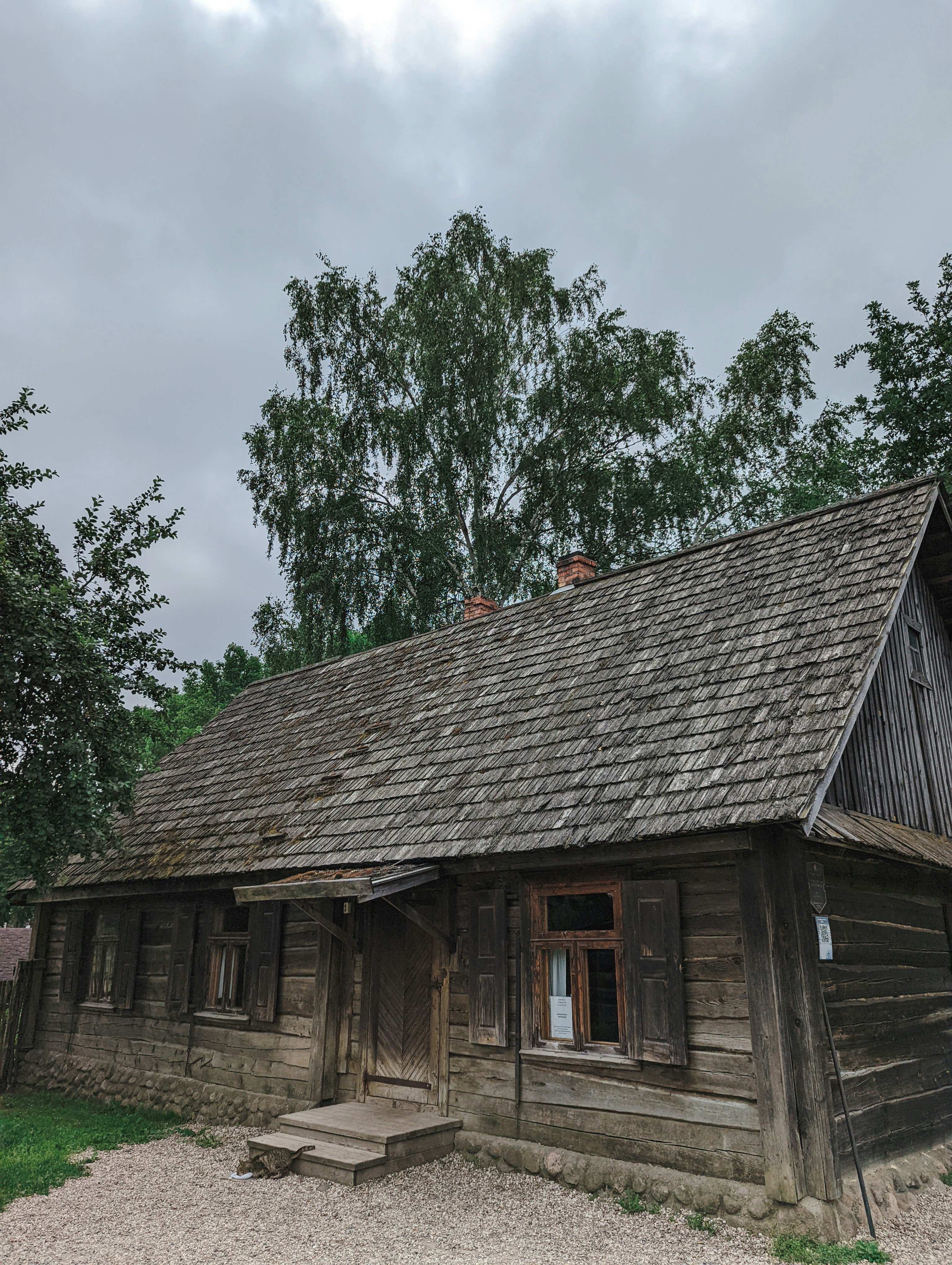 A rustic wooden house nestled among lush greenery, showcasing traditional architecture with a thatched roof under a cloudy sky.