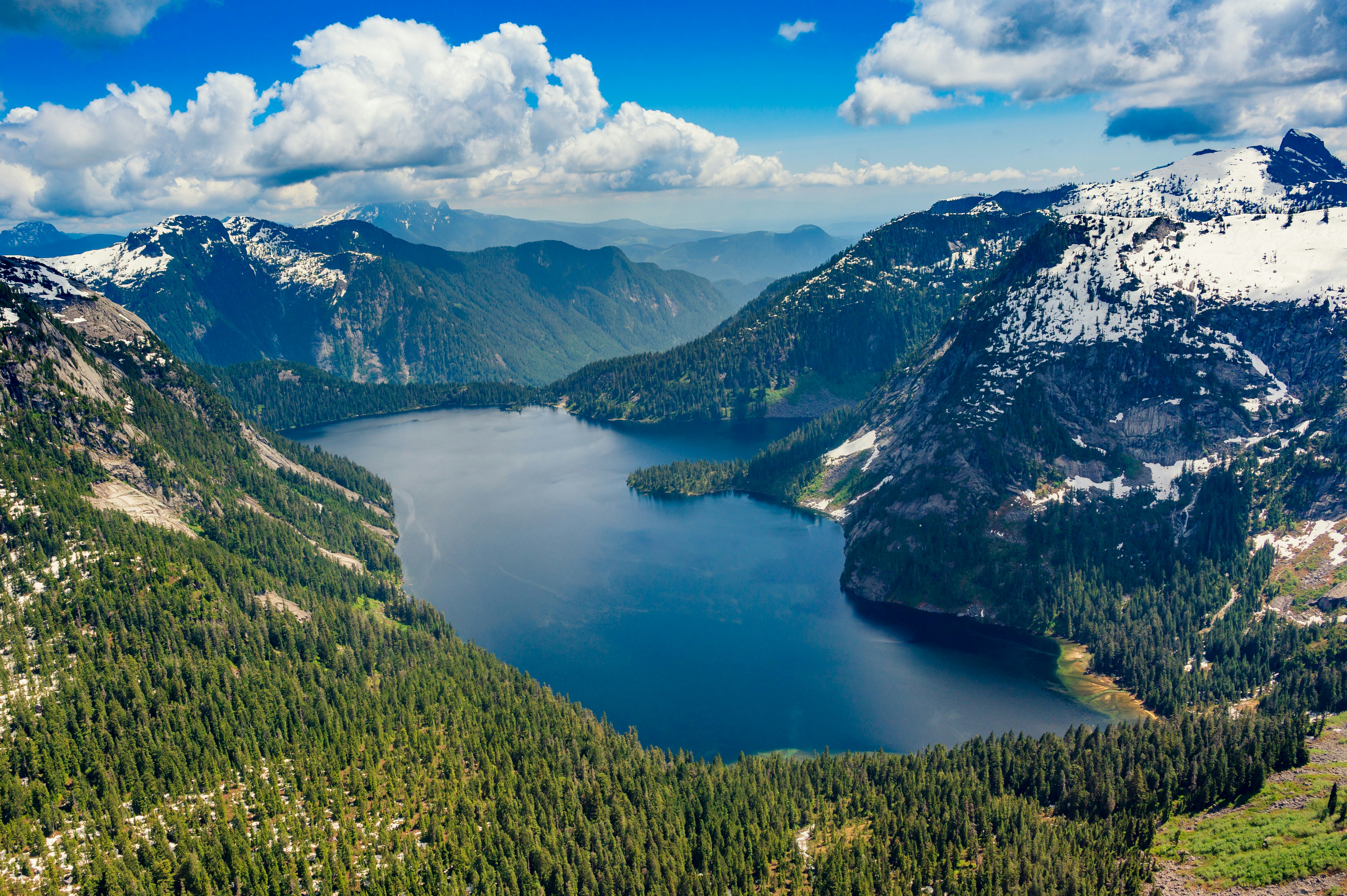 Aerial view of a tranquil lake surrounded by lush forests and snow-capped mountains under a partly cloudy sky.